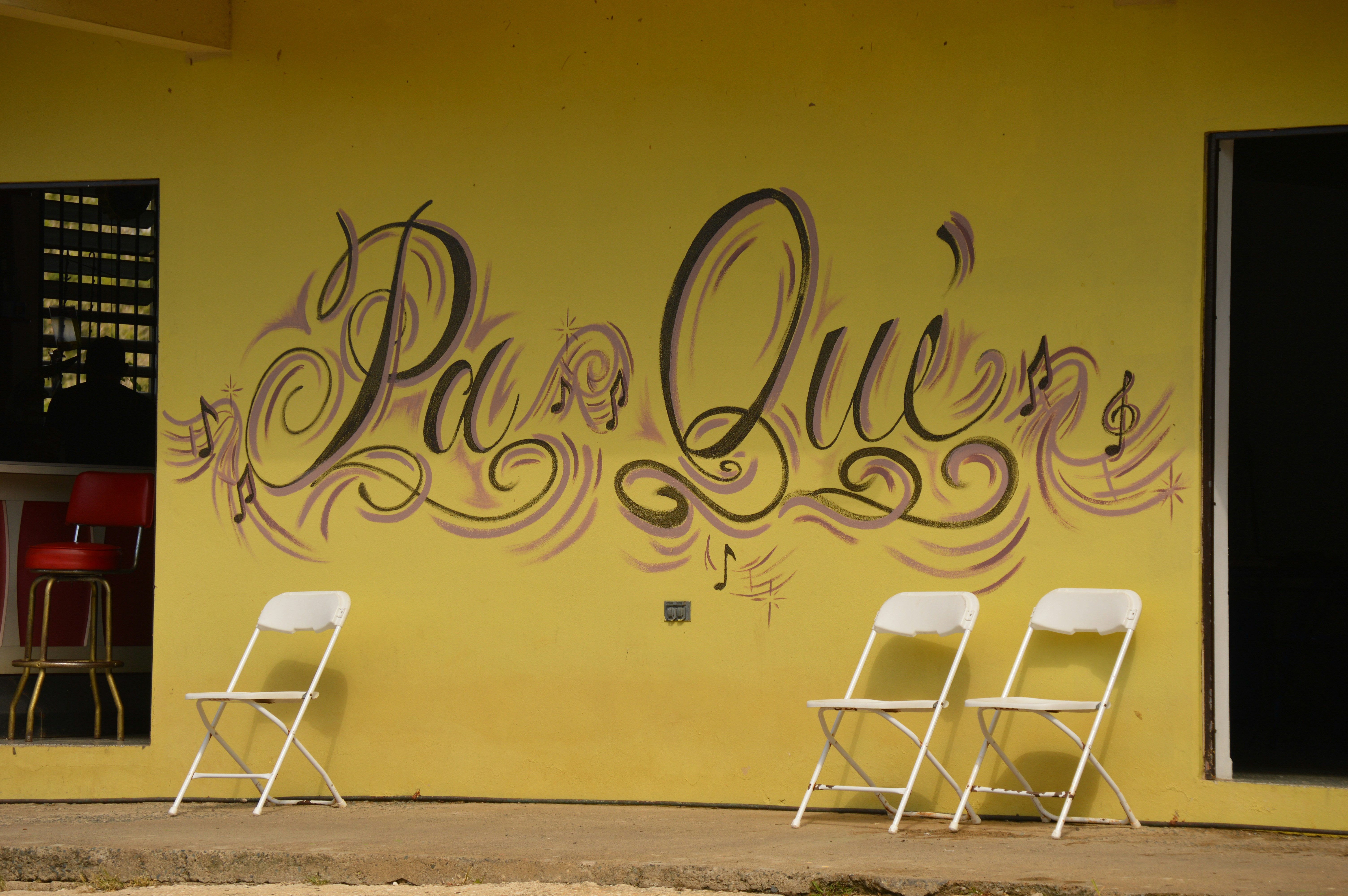 Ornate Graffiti on a Sunlit Yellow Wall with Idle White Chairs