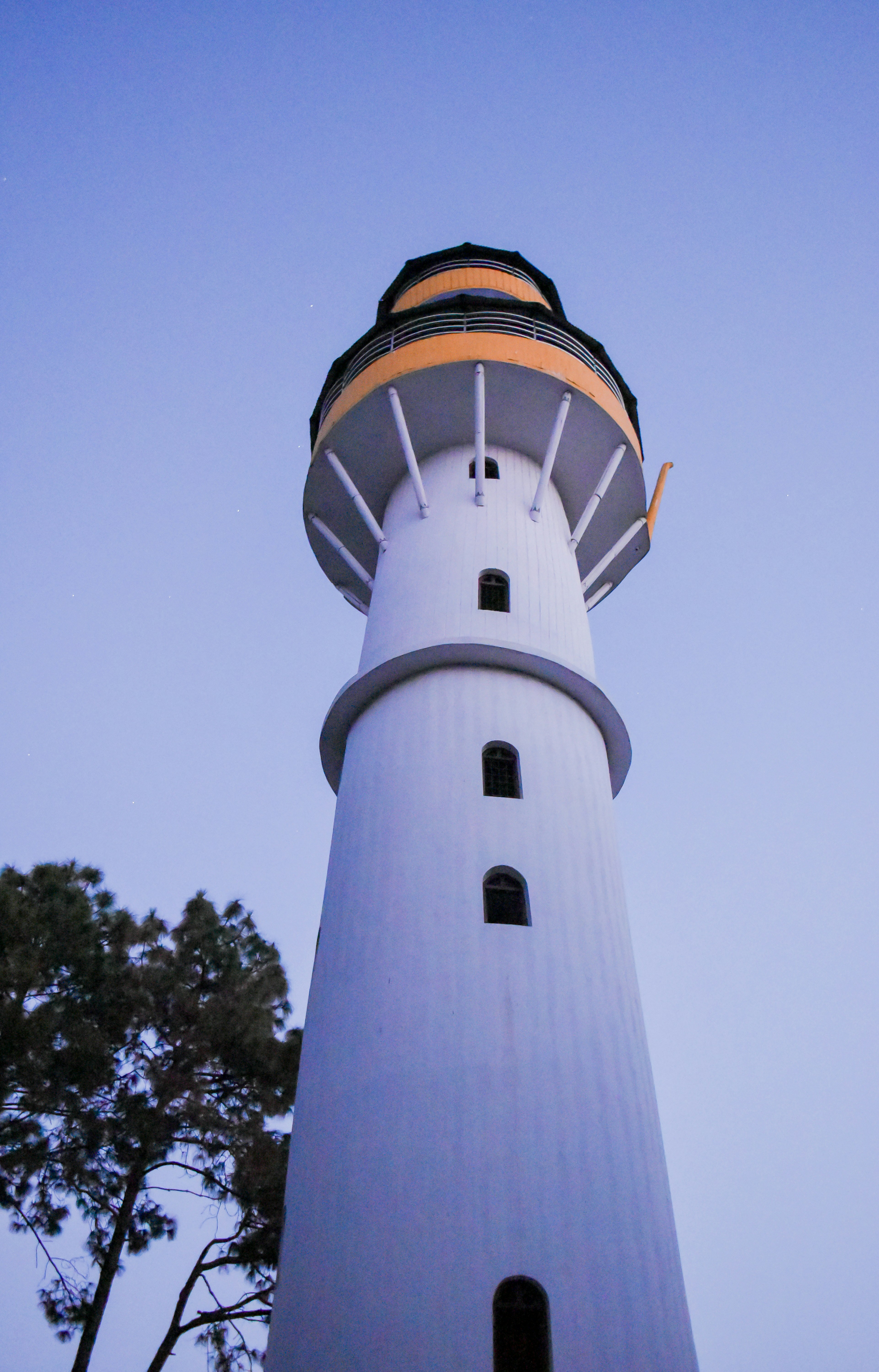 A tall lighthouse stands against a twilight sky, showcasing its white structure and golden accents. The silhouette of nearby trees adds depth to the serene scene.