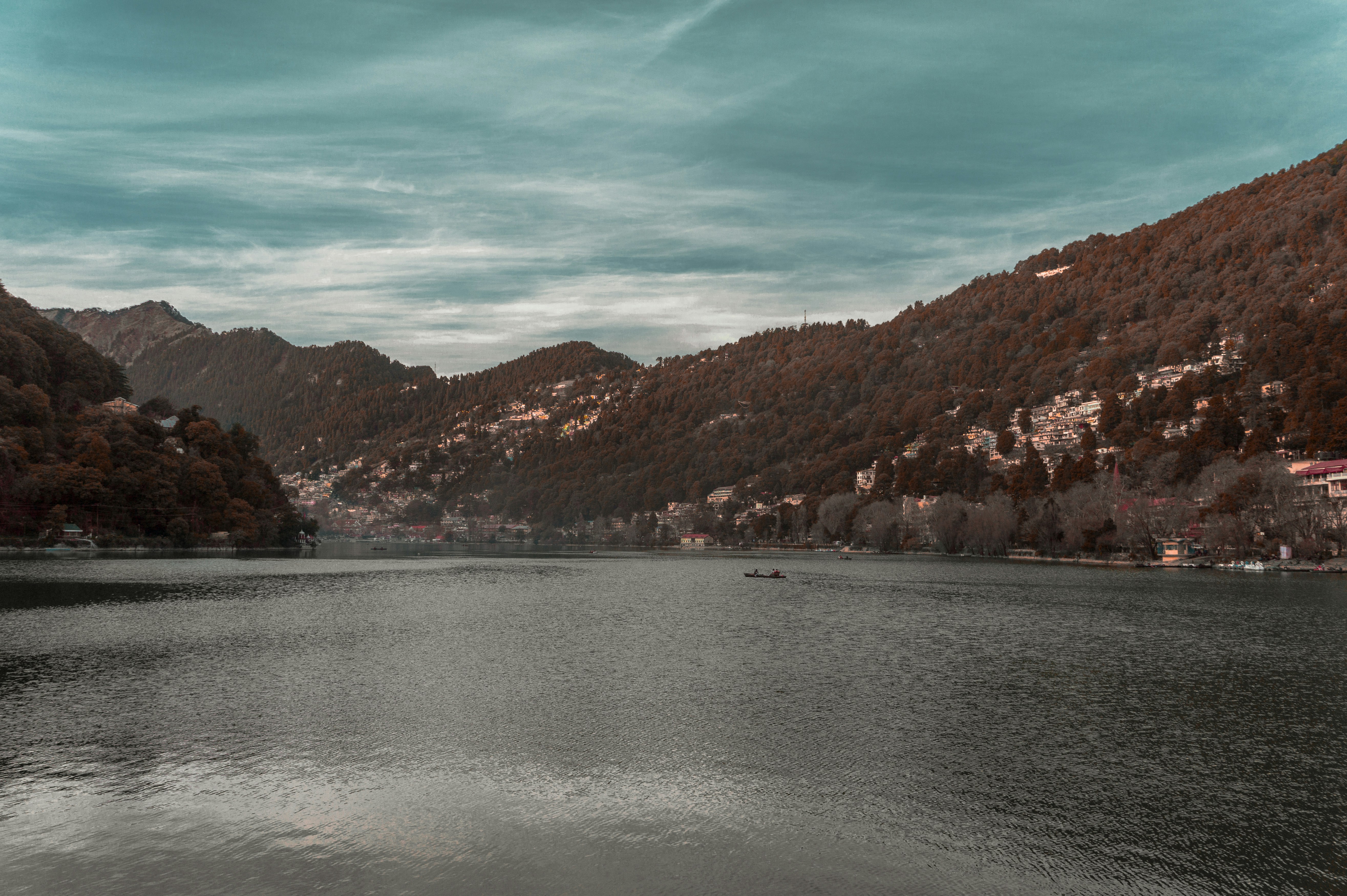 Photograph of a tranquil alpine lake framed by terracotta-hued mountains, with a distant shoreline dotted by houses under a teal-tinged sky.