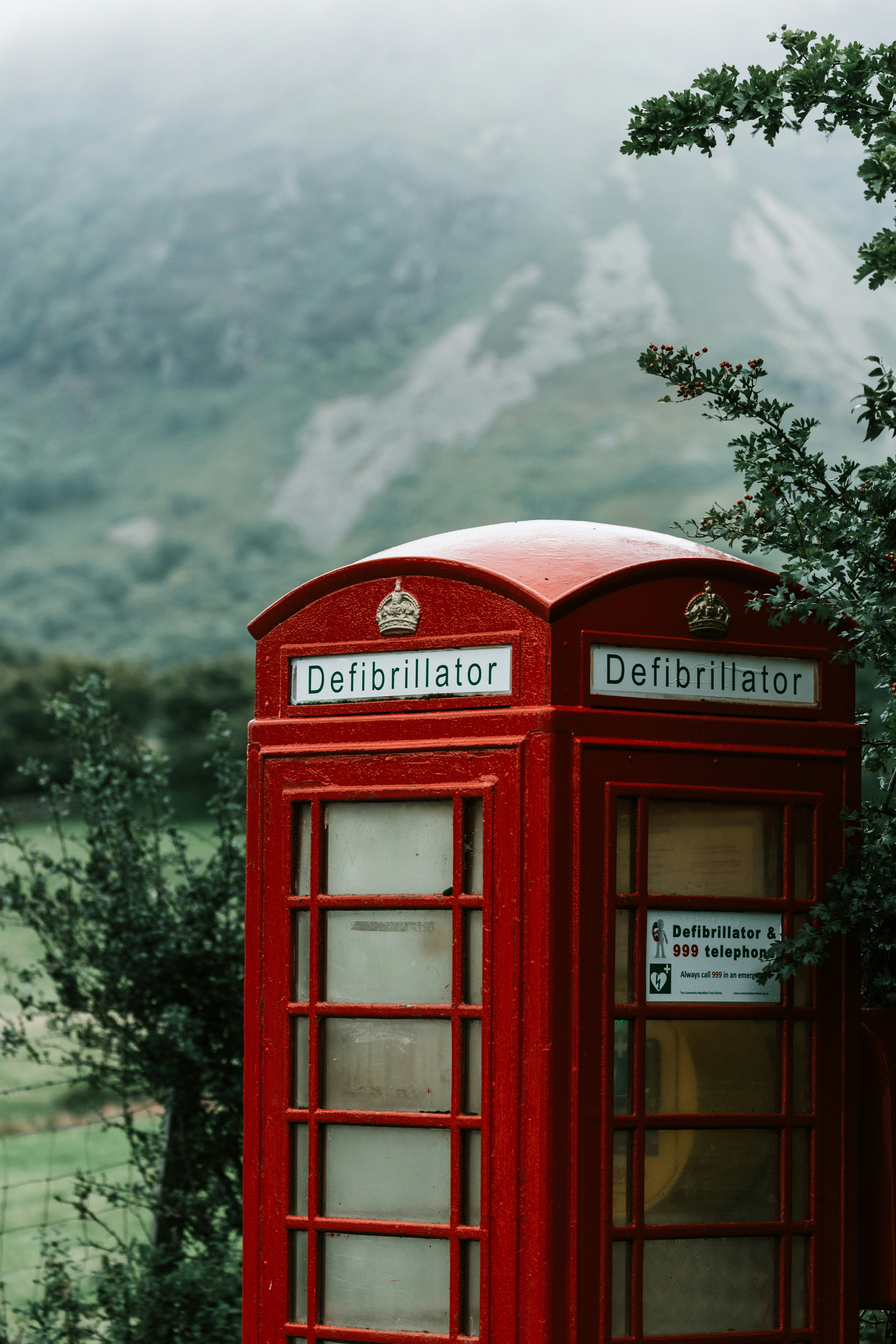 A vintage red telephone box repurposed as a defibrillator station, surrounded by lush greenery and misty hills in the background.