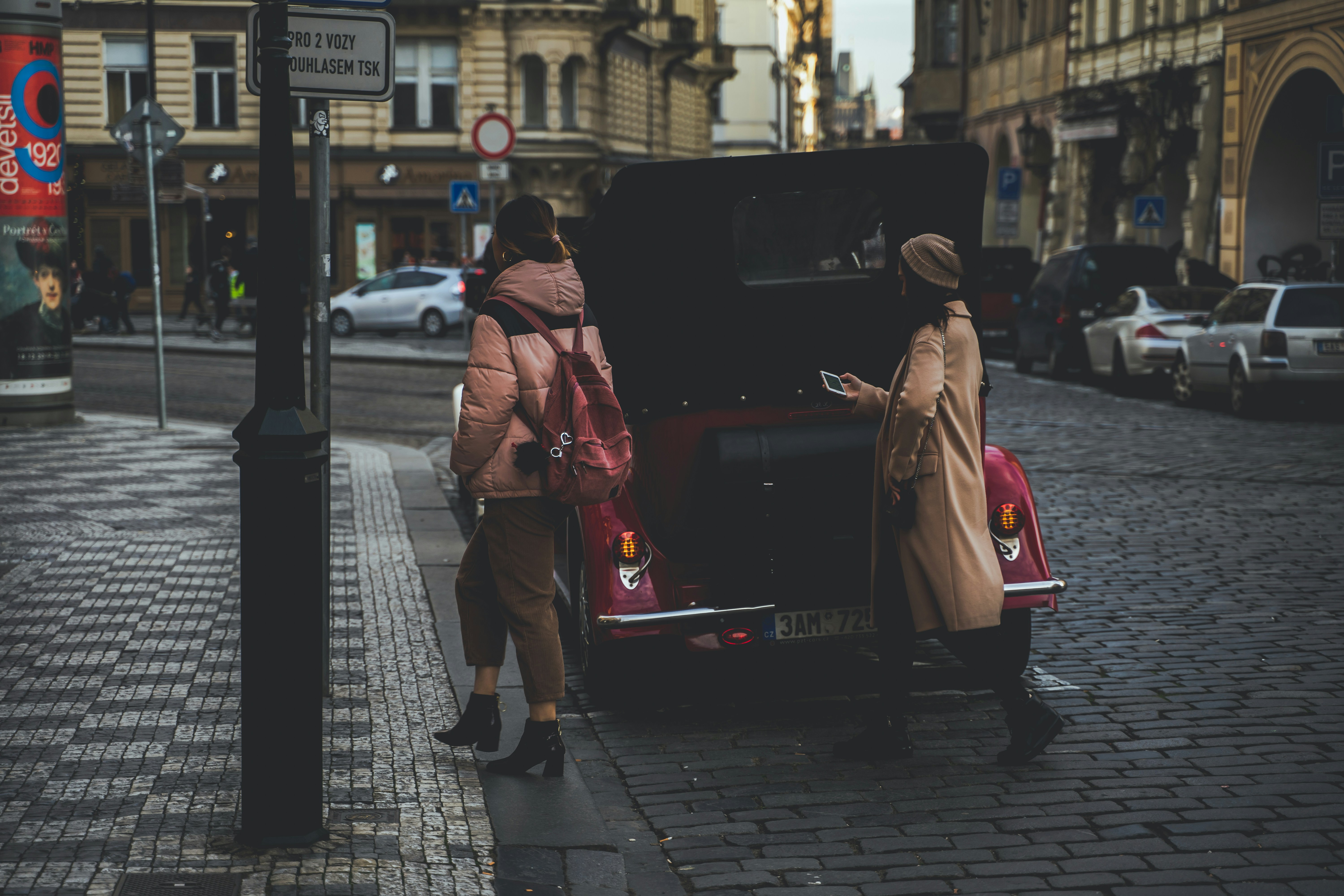 Two women in stylish winter attire walk past a vintage car parked on a cobblestone street, embodying a blend of modern fashion and classic charm.