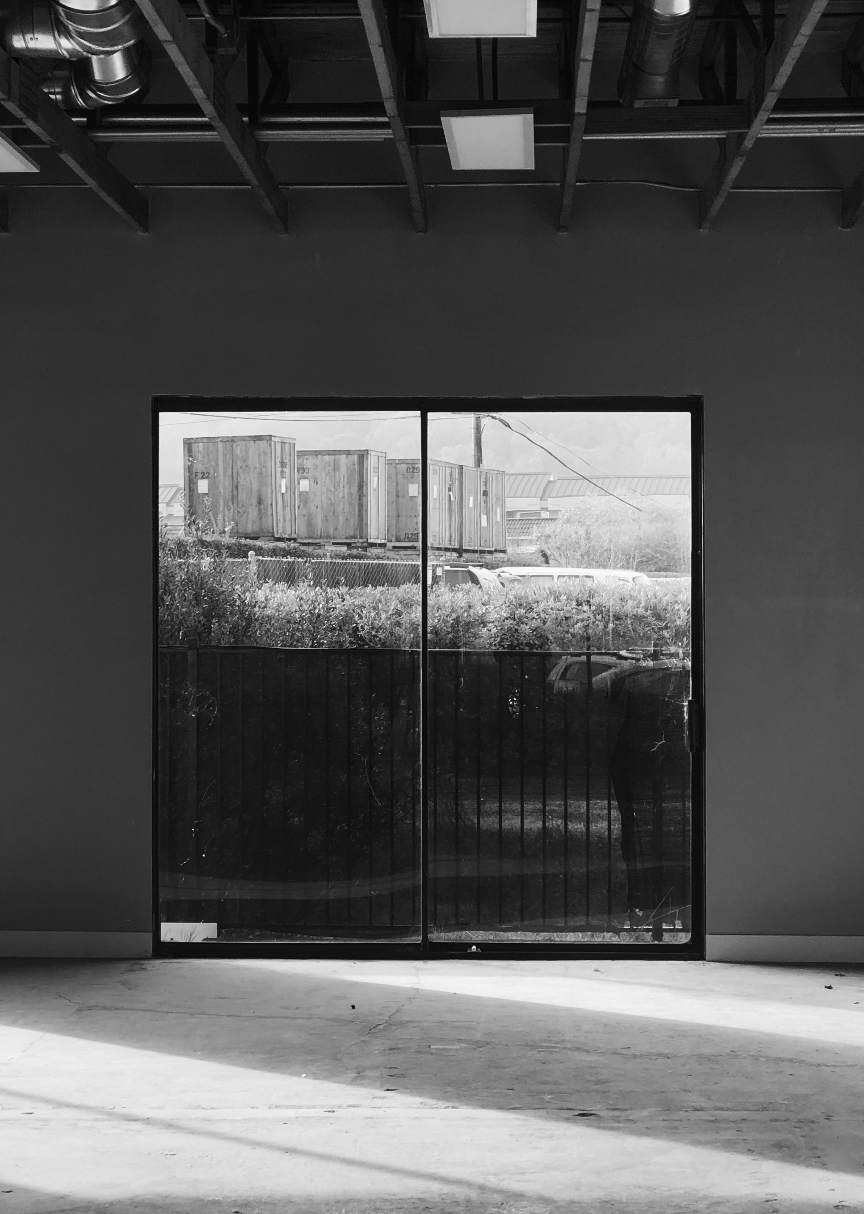 Black and white interior shot featuring a large window that frames wooden shipping containers and a distant landscape, emphasizing the contrast between inside and outside.
