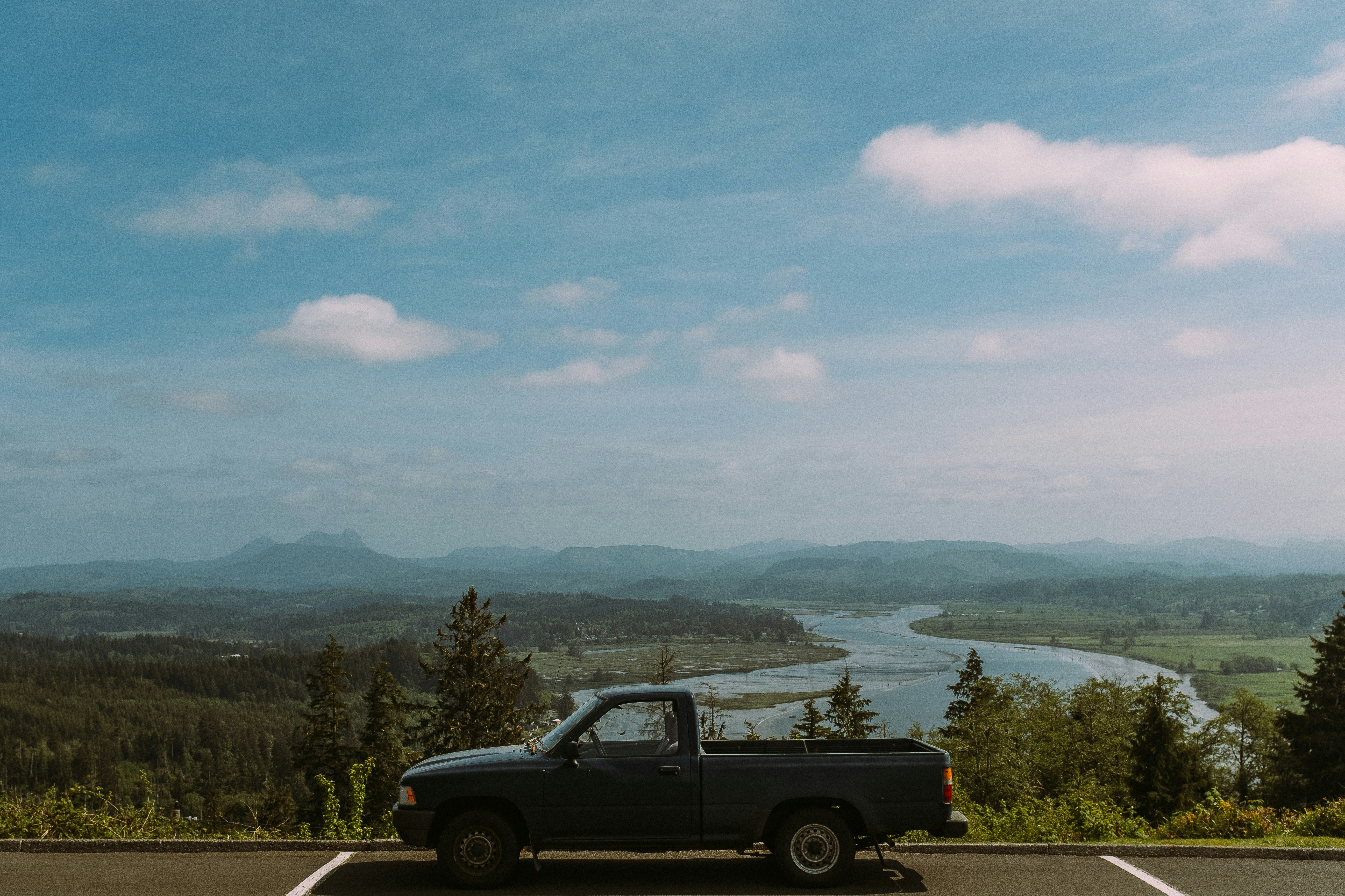 A vintage truck parked against a vast landscape with a winding river and distant mountains under a partly cloudy sky.