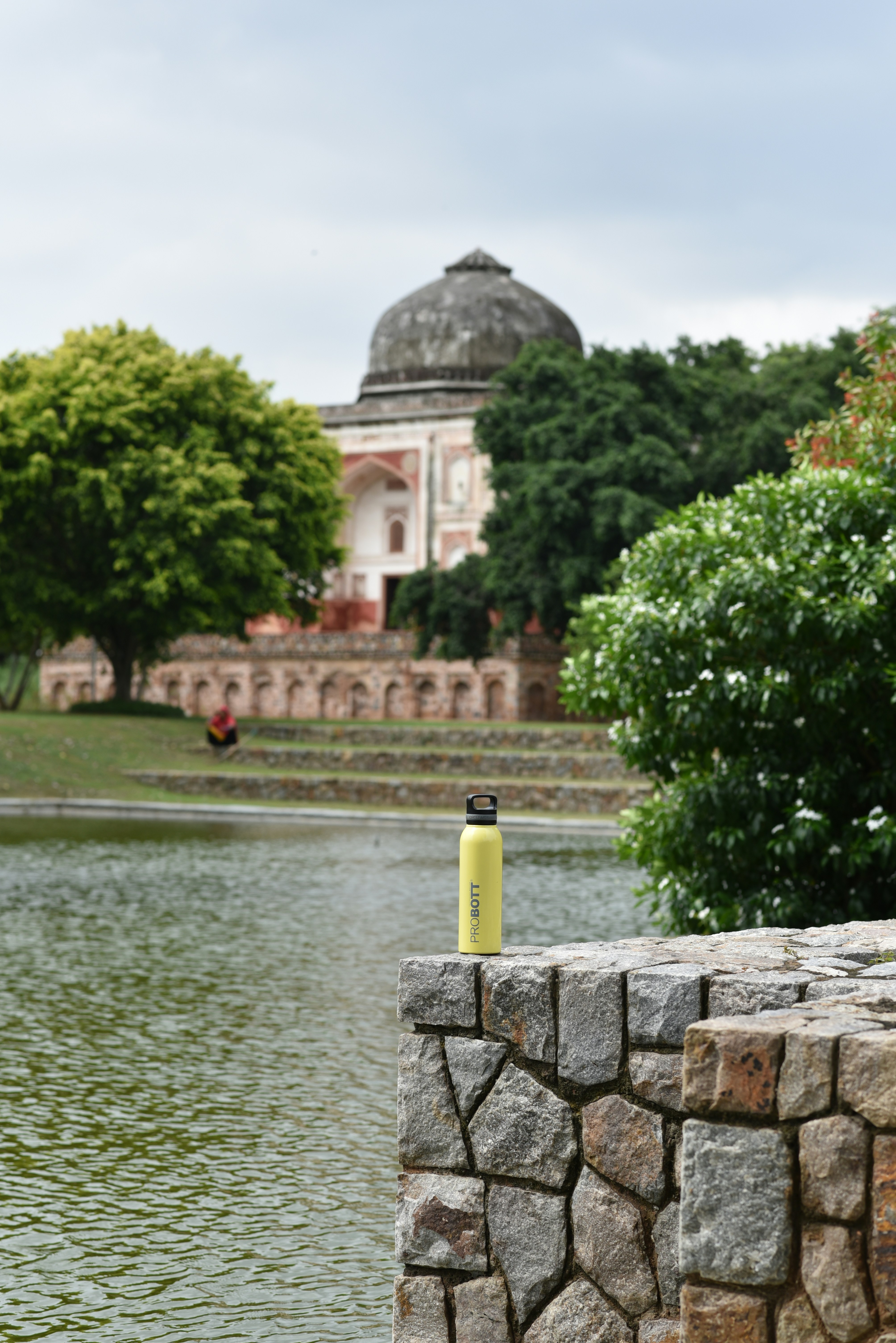 Yellow water bottle perched on a stone ledge beside a tranquil pond, with lush greenery and historical architecture in the background.