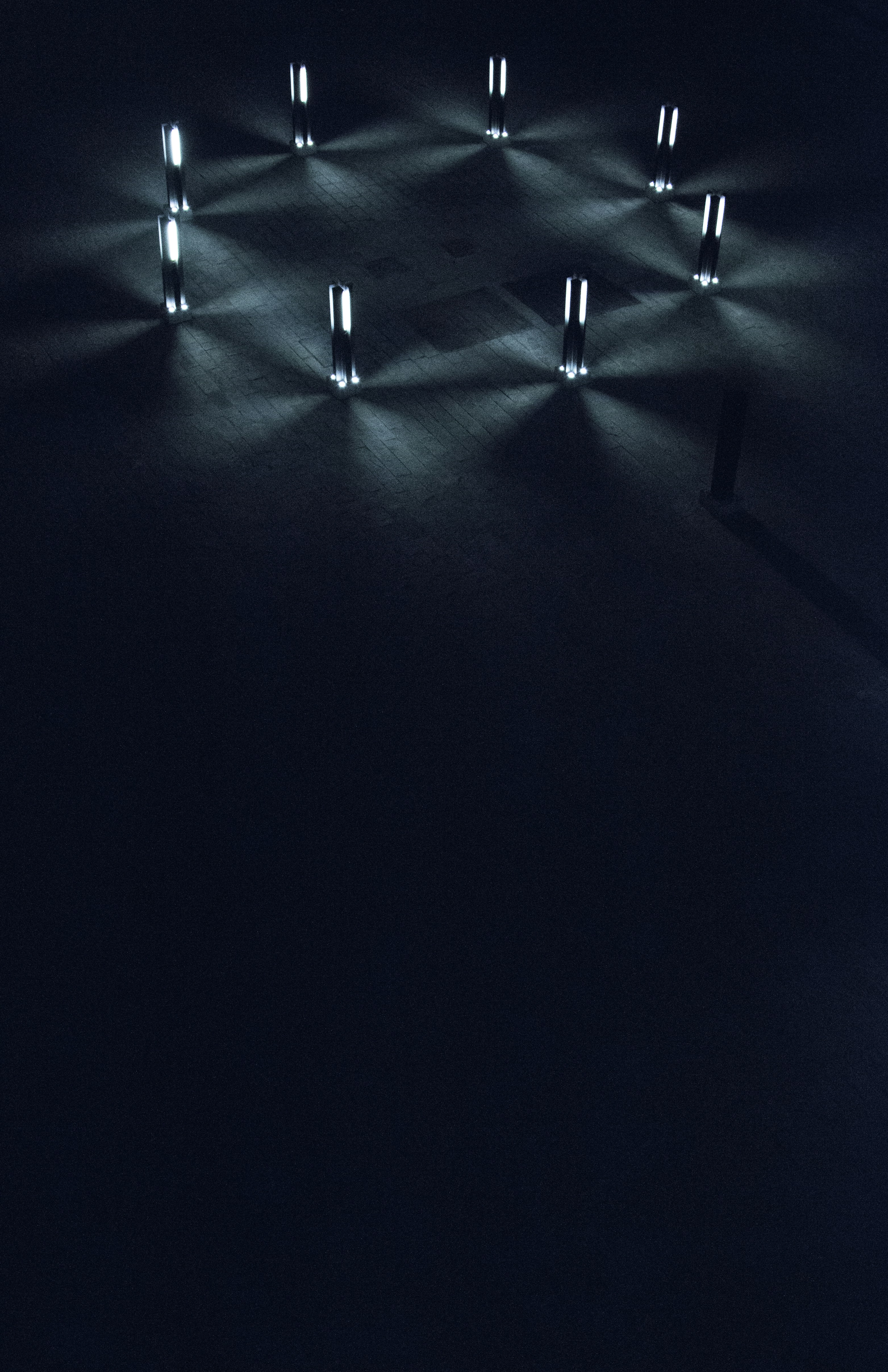 A circular arrangement of illuminated posts casting dramatic shadows on a dark surface, creating a geometric pattern. The scene evokes a sense of mystery and intrigue.