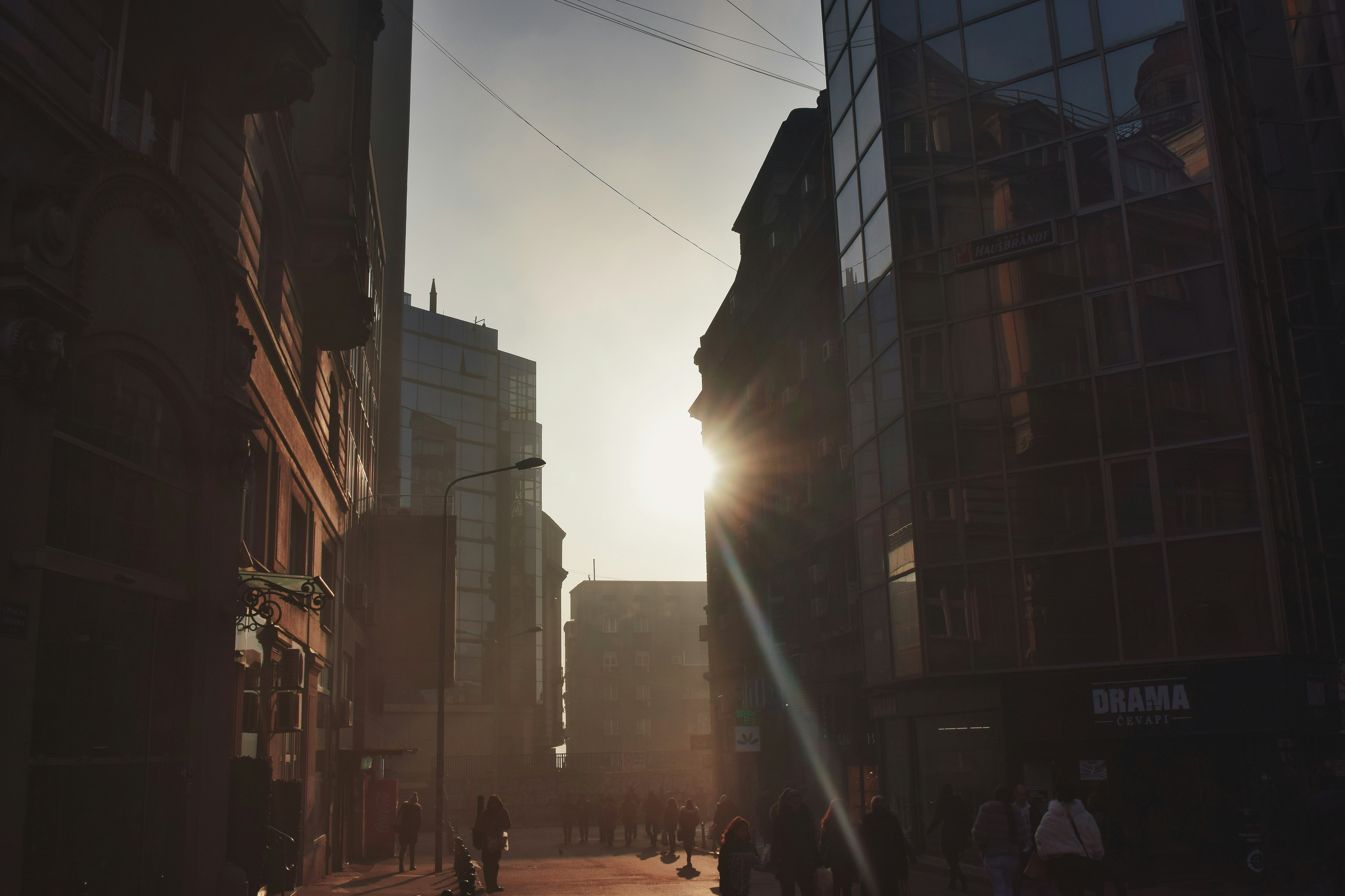 Pedestrians walk along a city street framed by tall buildings as the sun peeks through, casting long shadows.