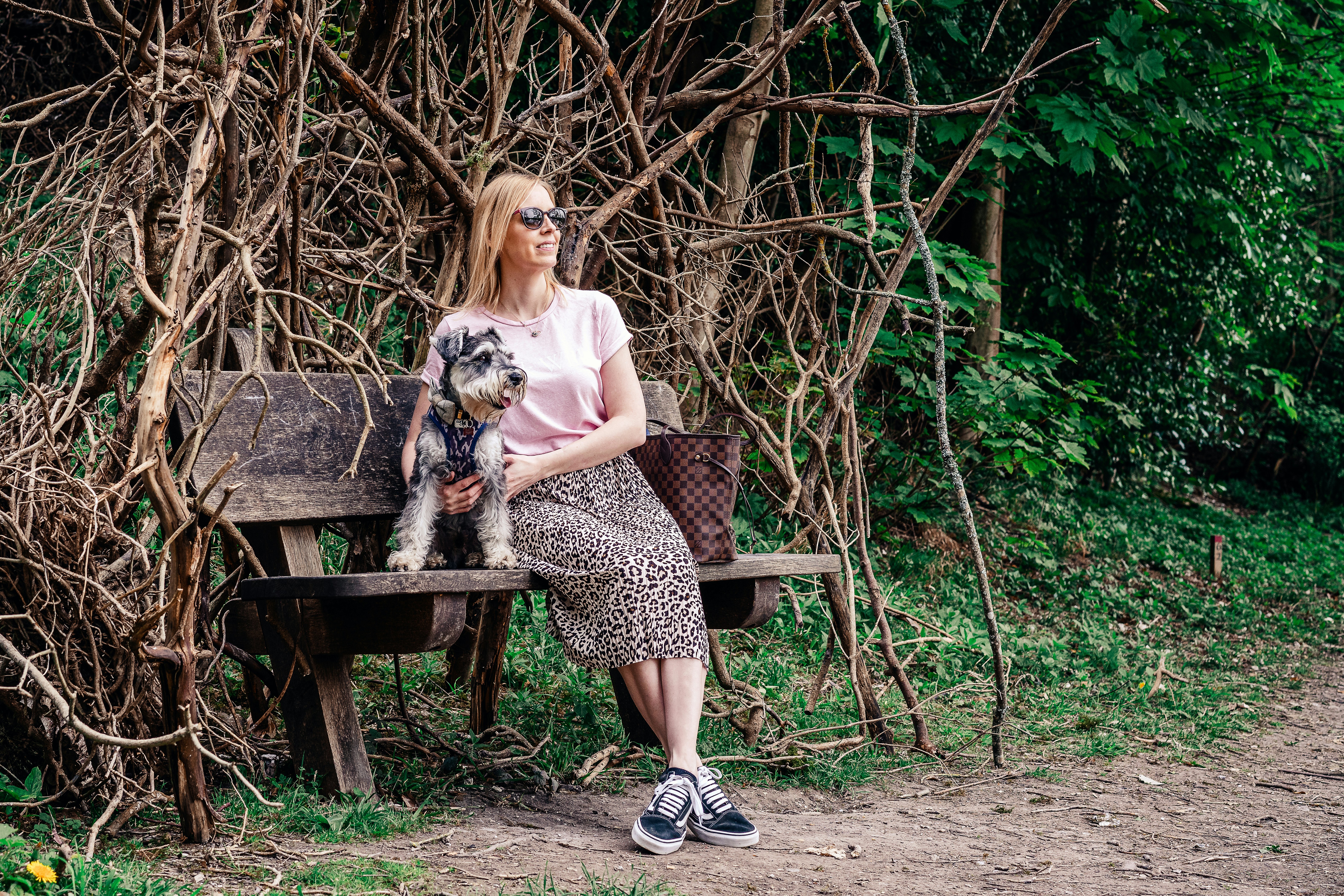 Woman seated on a bench with her dog, surrounded by intertwining branches and lush greenery.