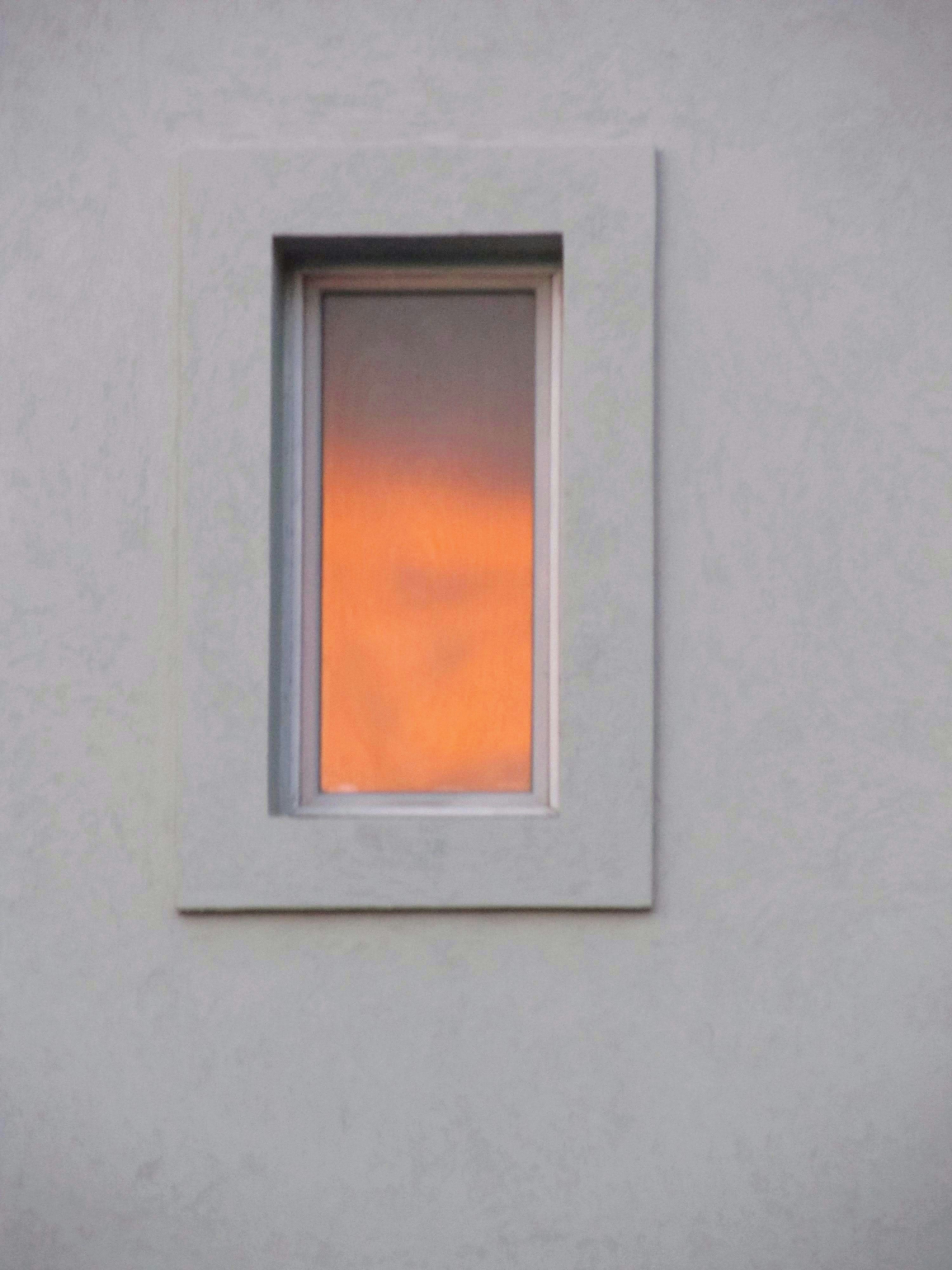 A slender vertical window in a pale stucco wall reflects a warm orange sunset glow.