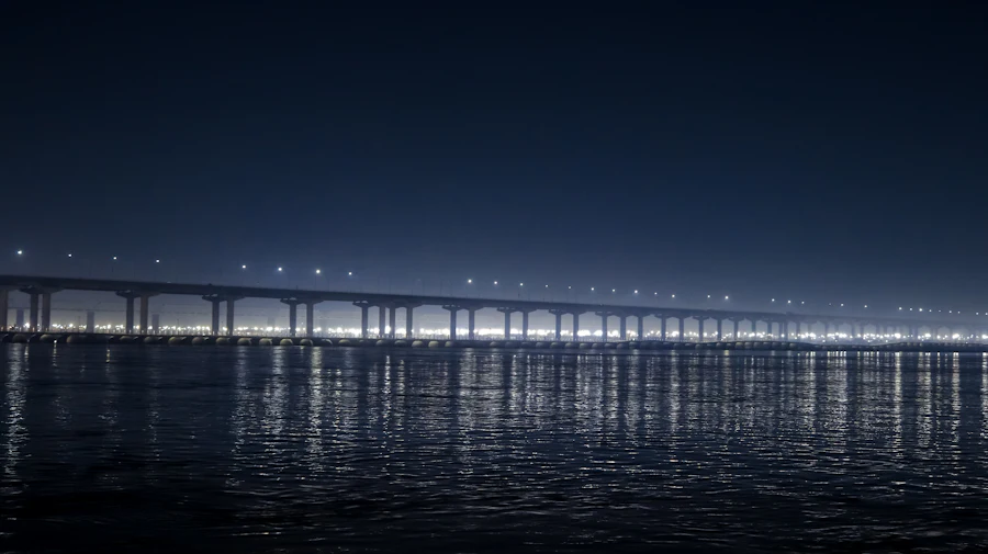A long, lit bridge spans across calm water at night, with city lights glowing in the distance and reflections shimmering on the surface.