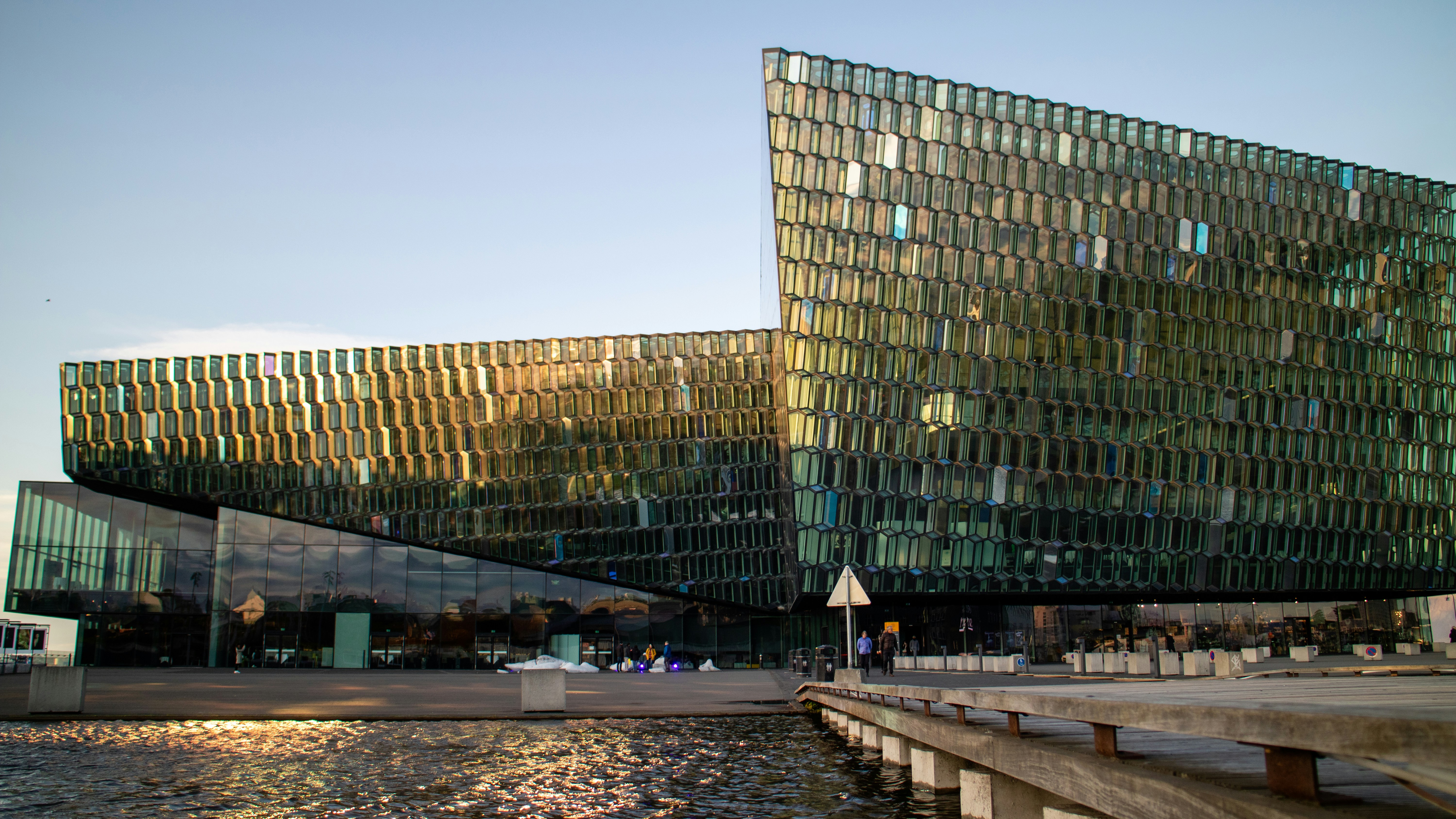 Modern glass facade of Harpa Concert Hall reflecting golden sunset hues, with melting icebergs near the entrance.