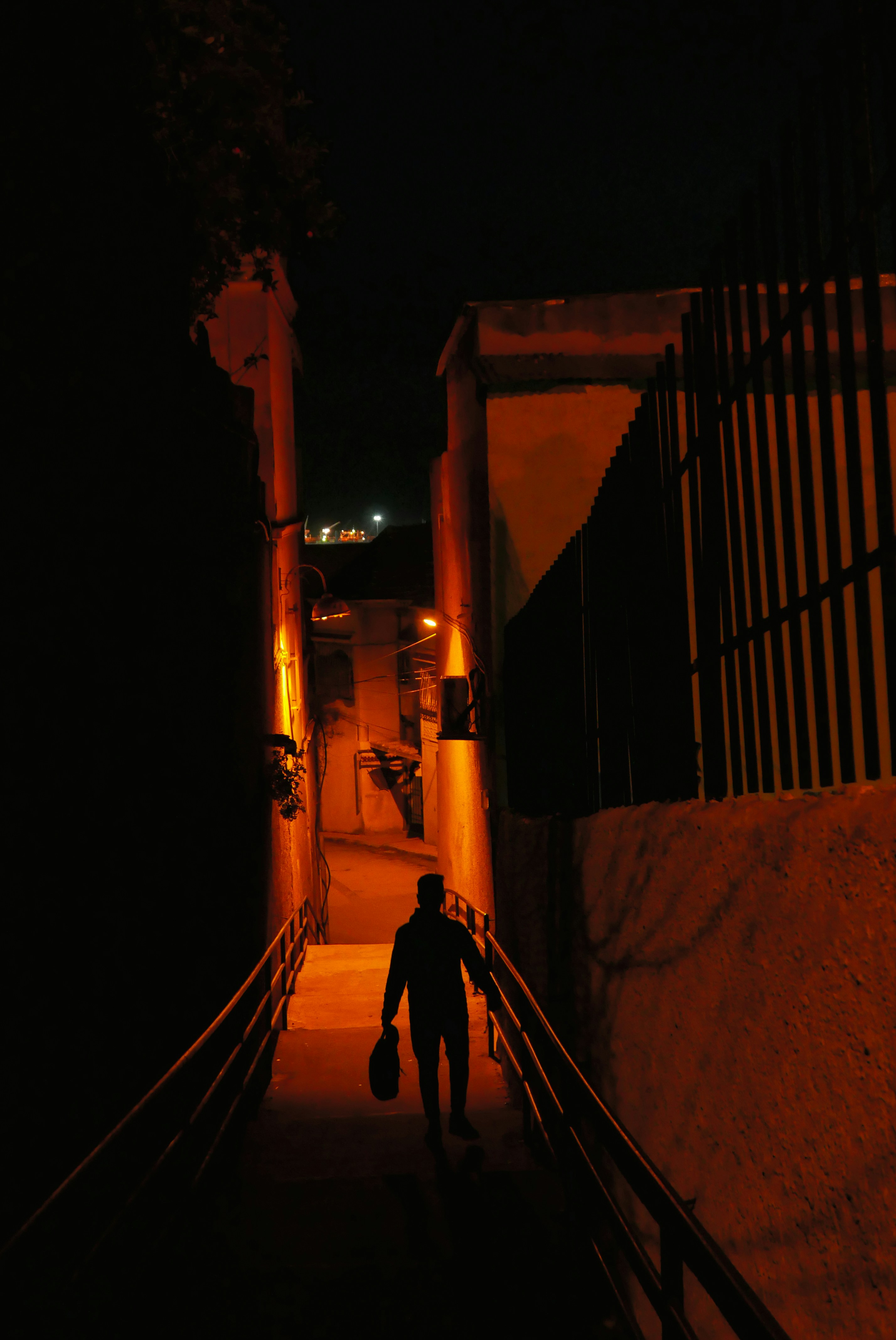 Silhouette of a person walking down a dimly lit alley, illuminated by warm orange streetlights. The scene evokes a sense of solitude and mystery.