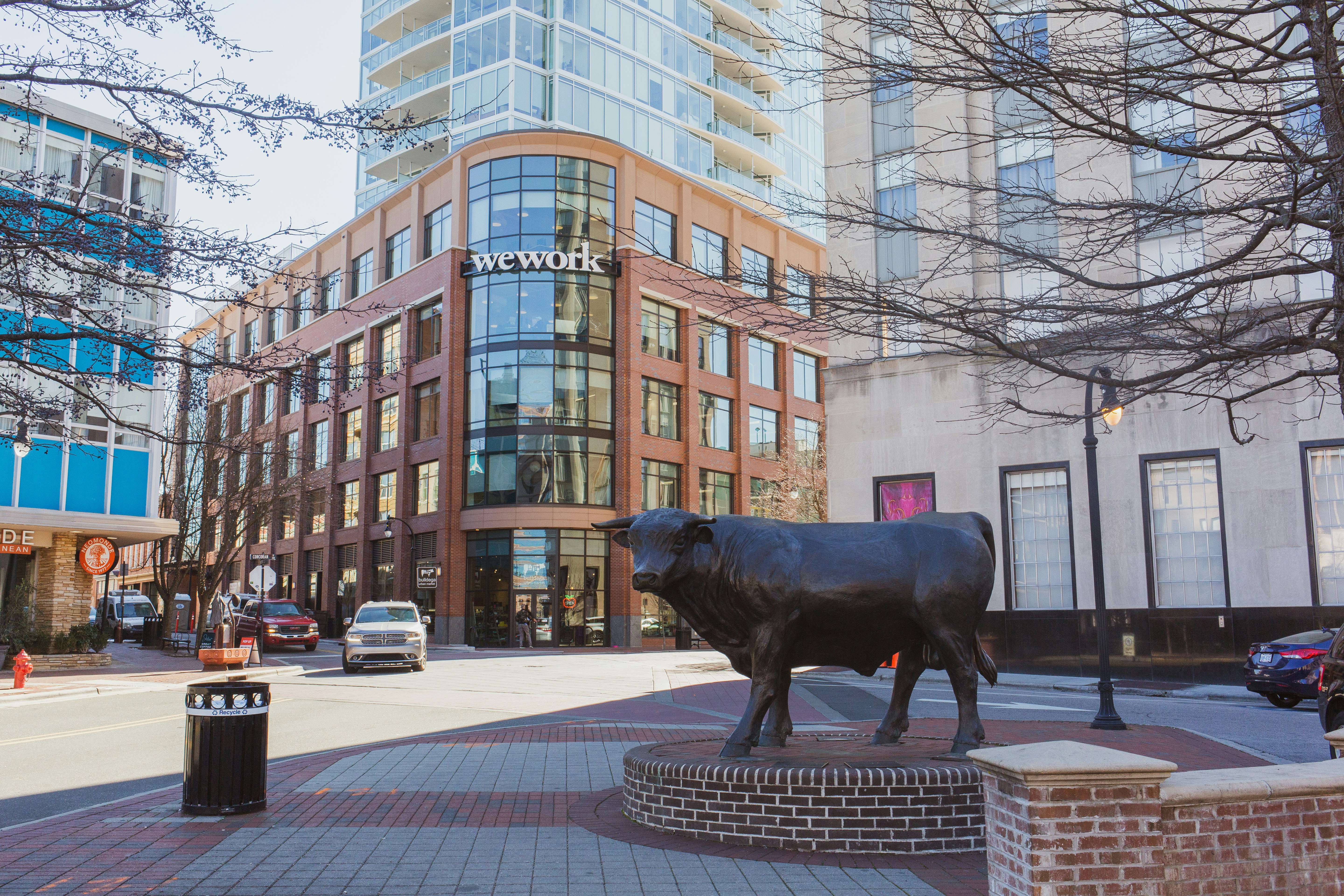 Bronze cow statue on a street corner amid modern city buildings during the day.