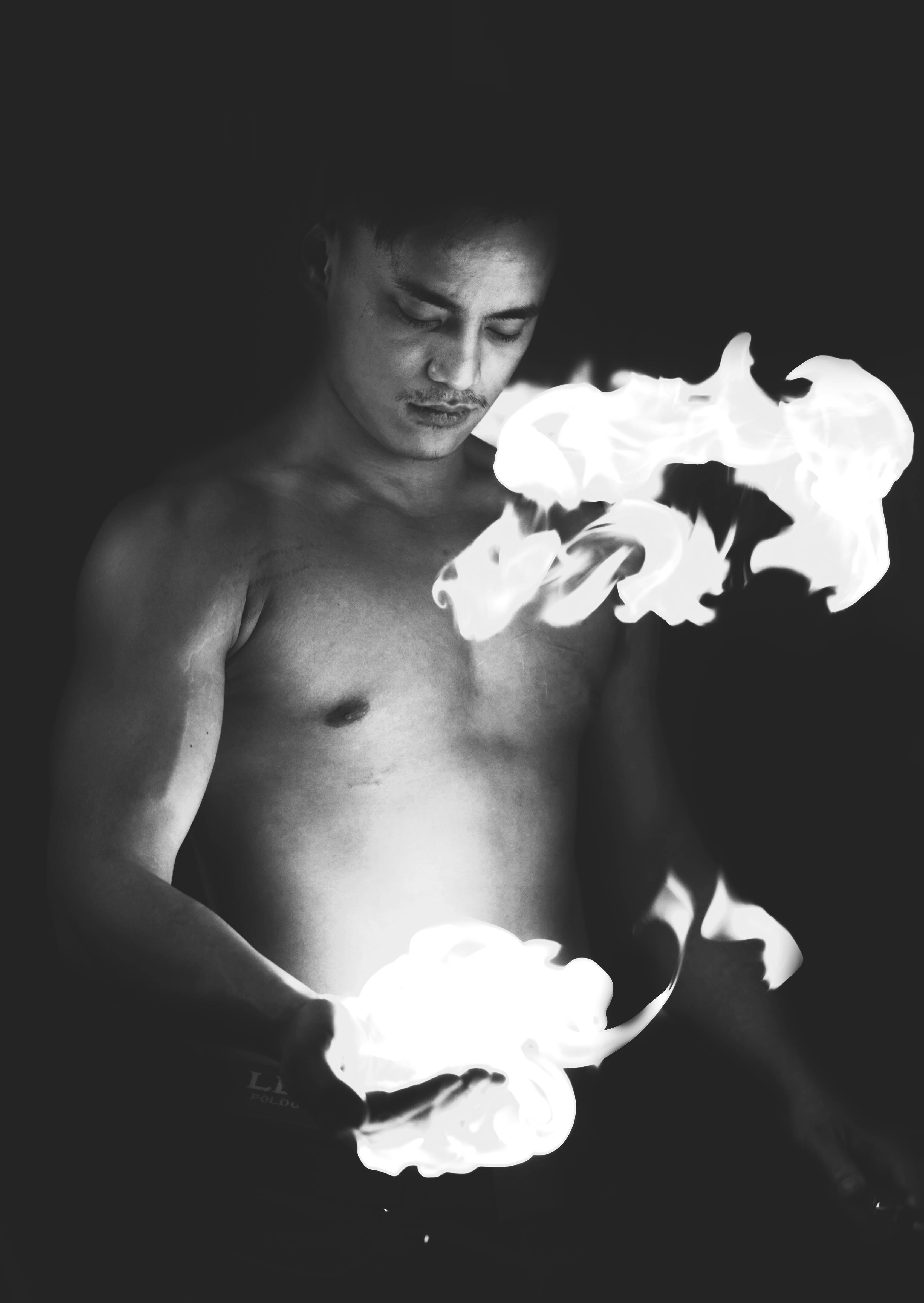 Black-and-white photograph of a shirtless man performing fire smoke manipulation, with bright wisps around his hands against a dark background.