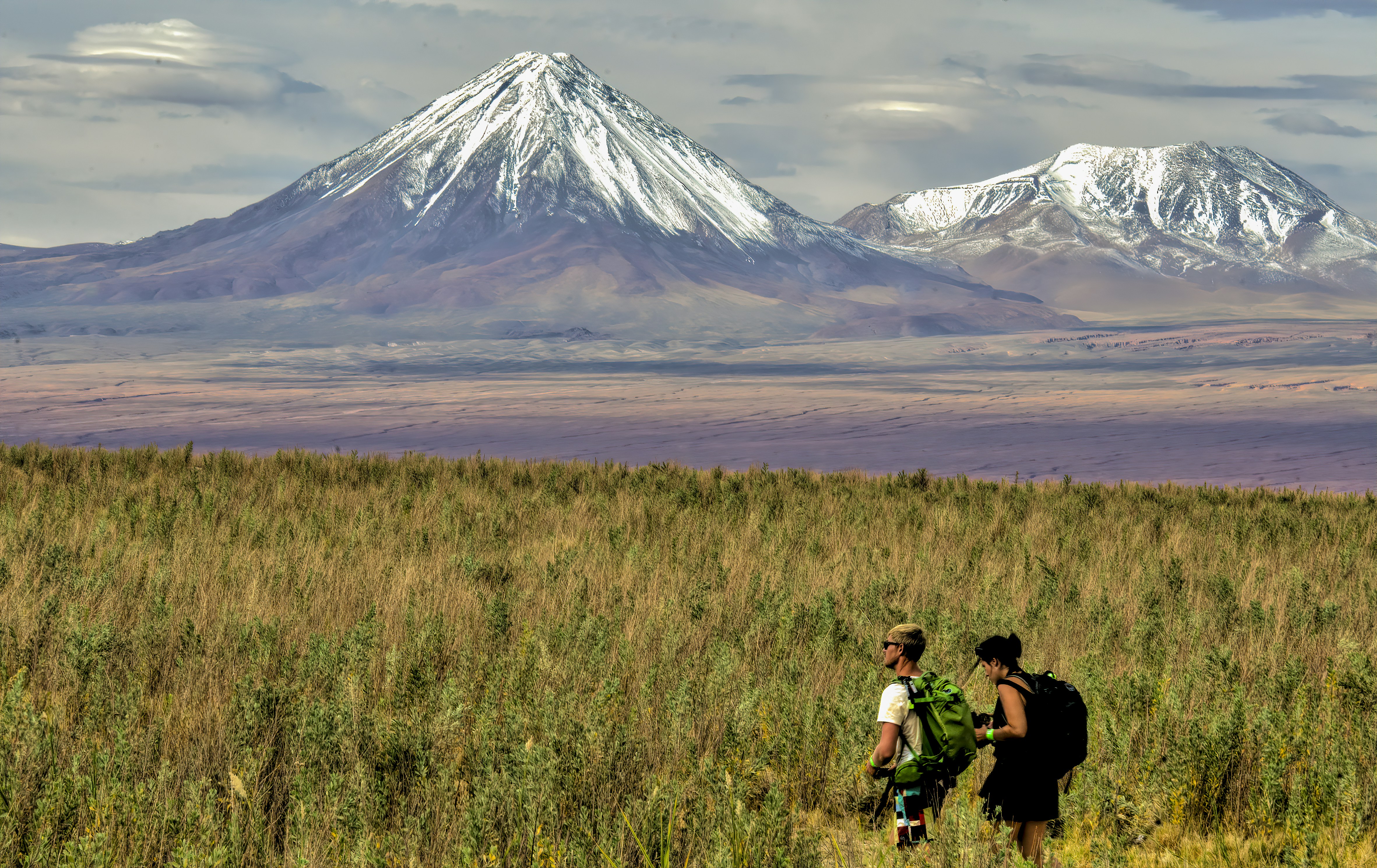 Couple hiking through grassy plains with snow-capped Andean peaks in the distance.