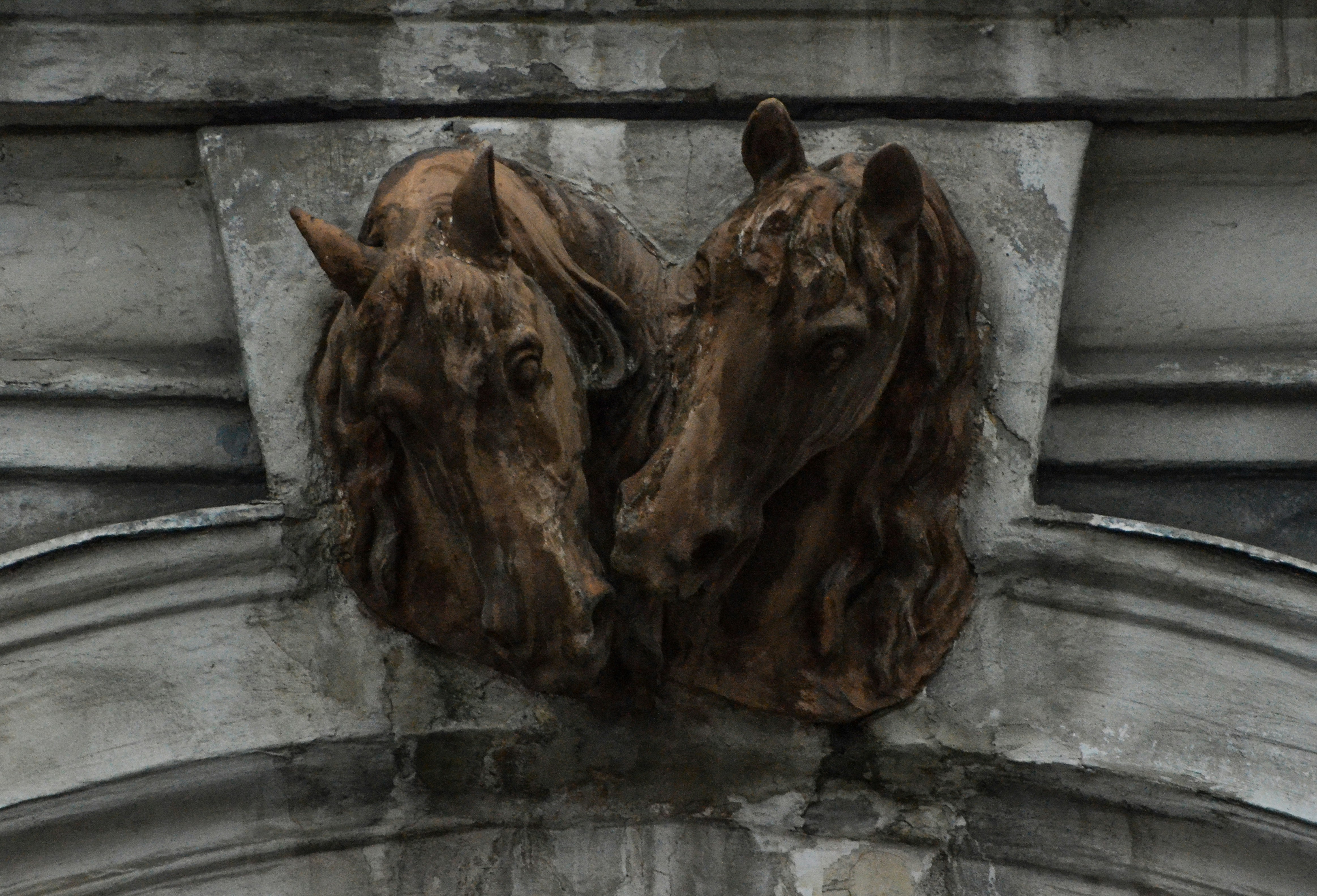 Two intricately carved horse heads merge in a sculptural display atop an architectural feature, showcasing artistry and detail. The weathered texture adds a sense of history.