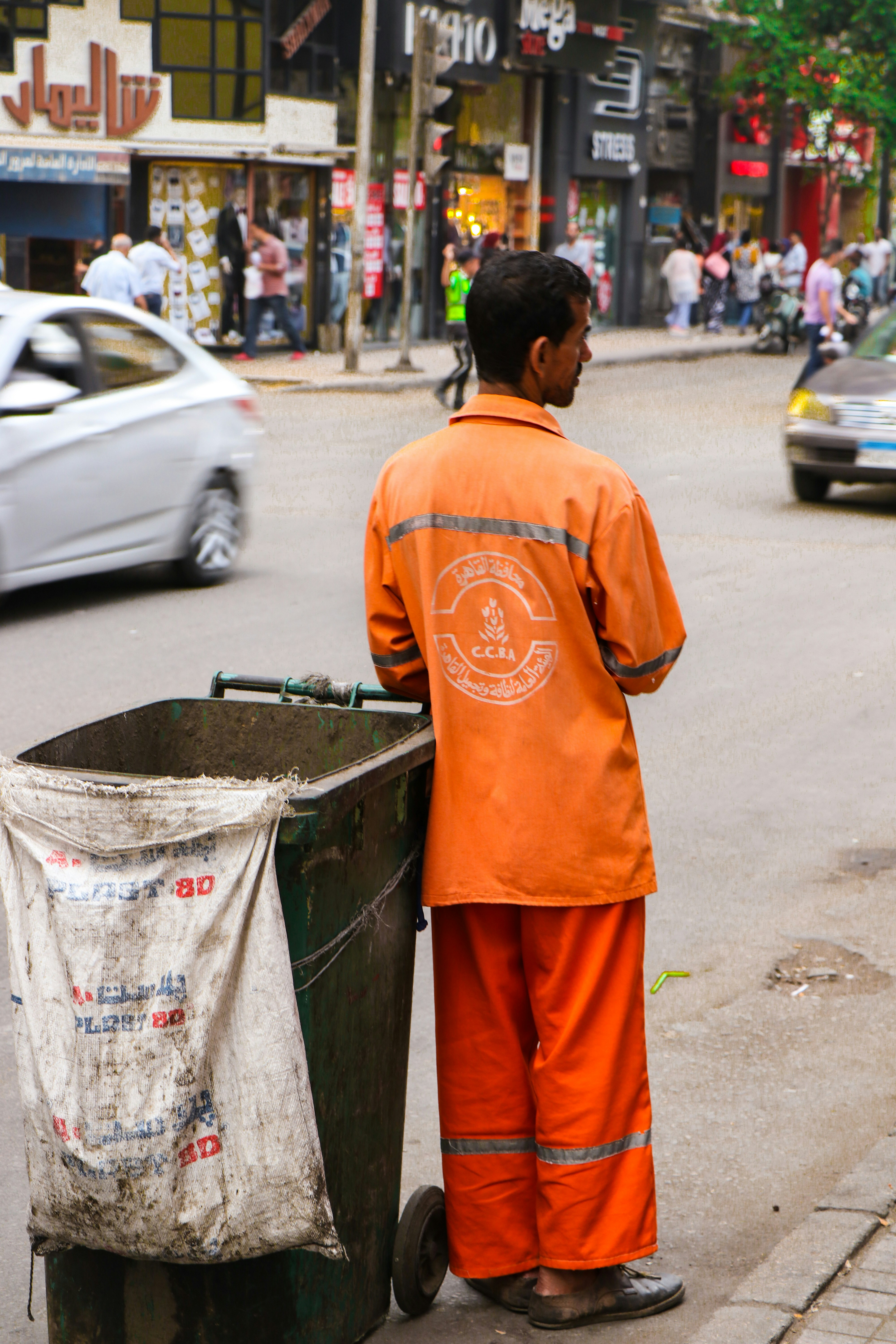 A sanitation worker in bright orange attire stands beside a green garbage cart, observing the bustling city traffic. The scene captures the contrast between his stillness and the movement around him.