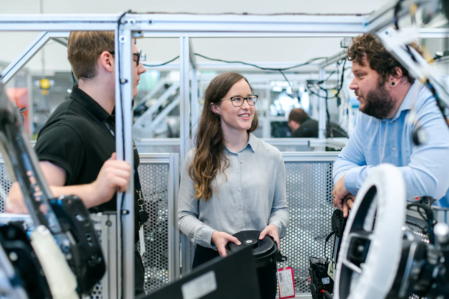 Photograph of three coworkers in a robotics assembly area, with the central woman engaging the group amid exposed machinery.
