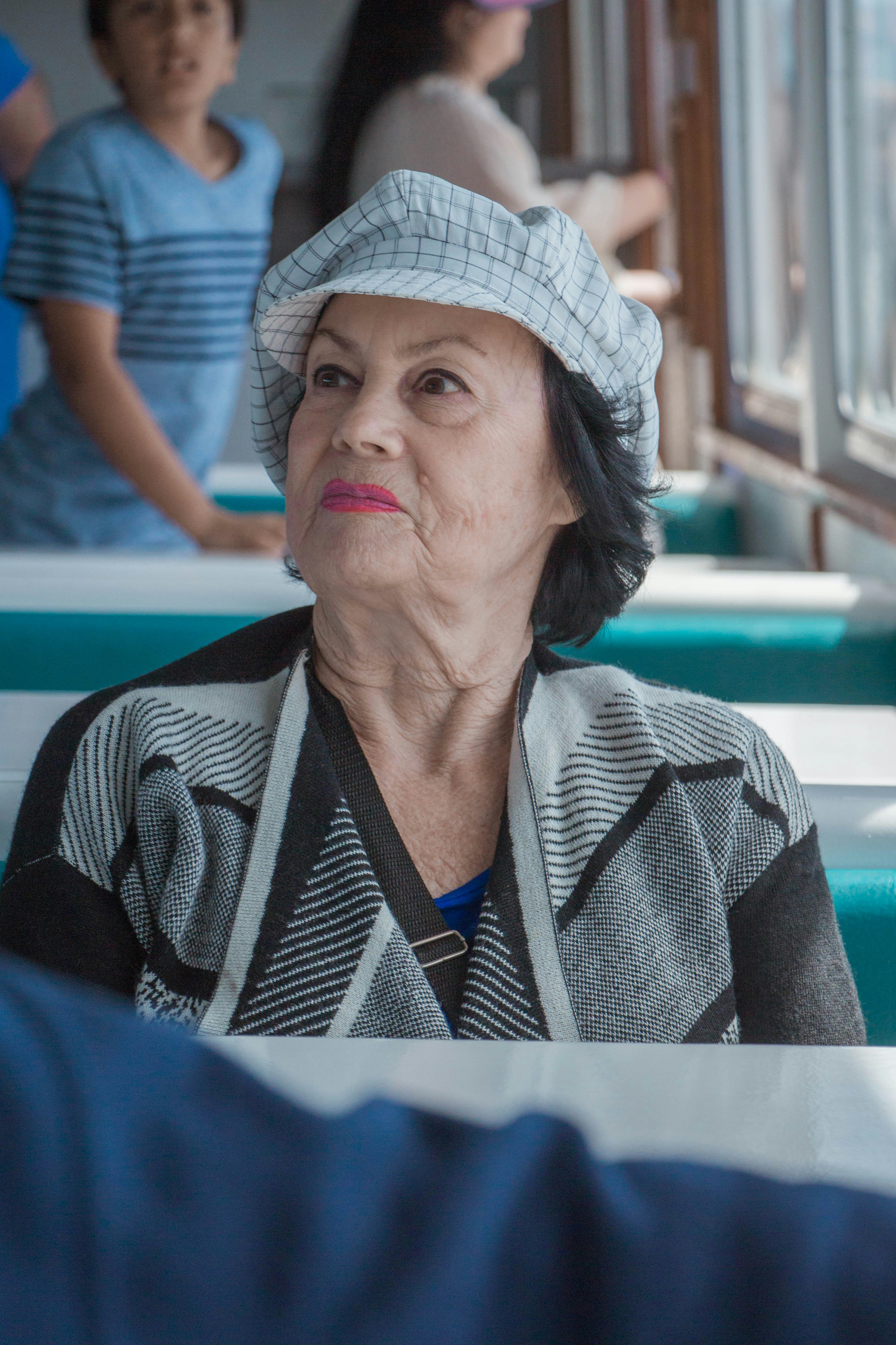Elderly woman with a thoughtful expression sits in a boat, surrounded by teal seating and natural light from nearby windows.
