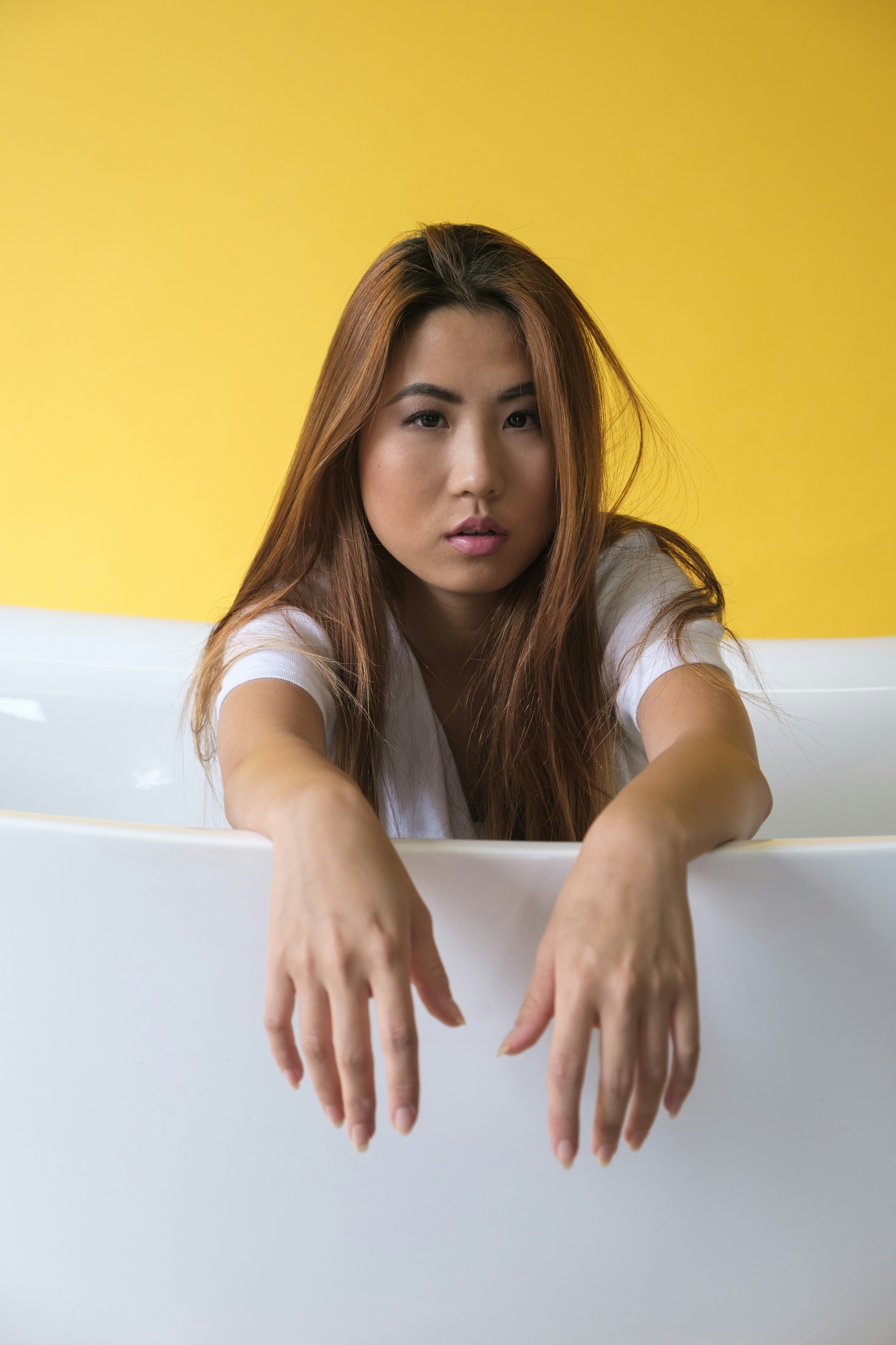 A young woman with long hair lounges in a white tub against a vibrant yellow backdrop, exuding a sense of calm and introspection.