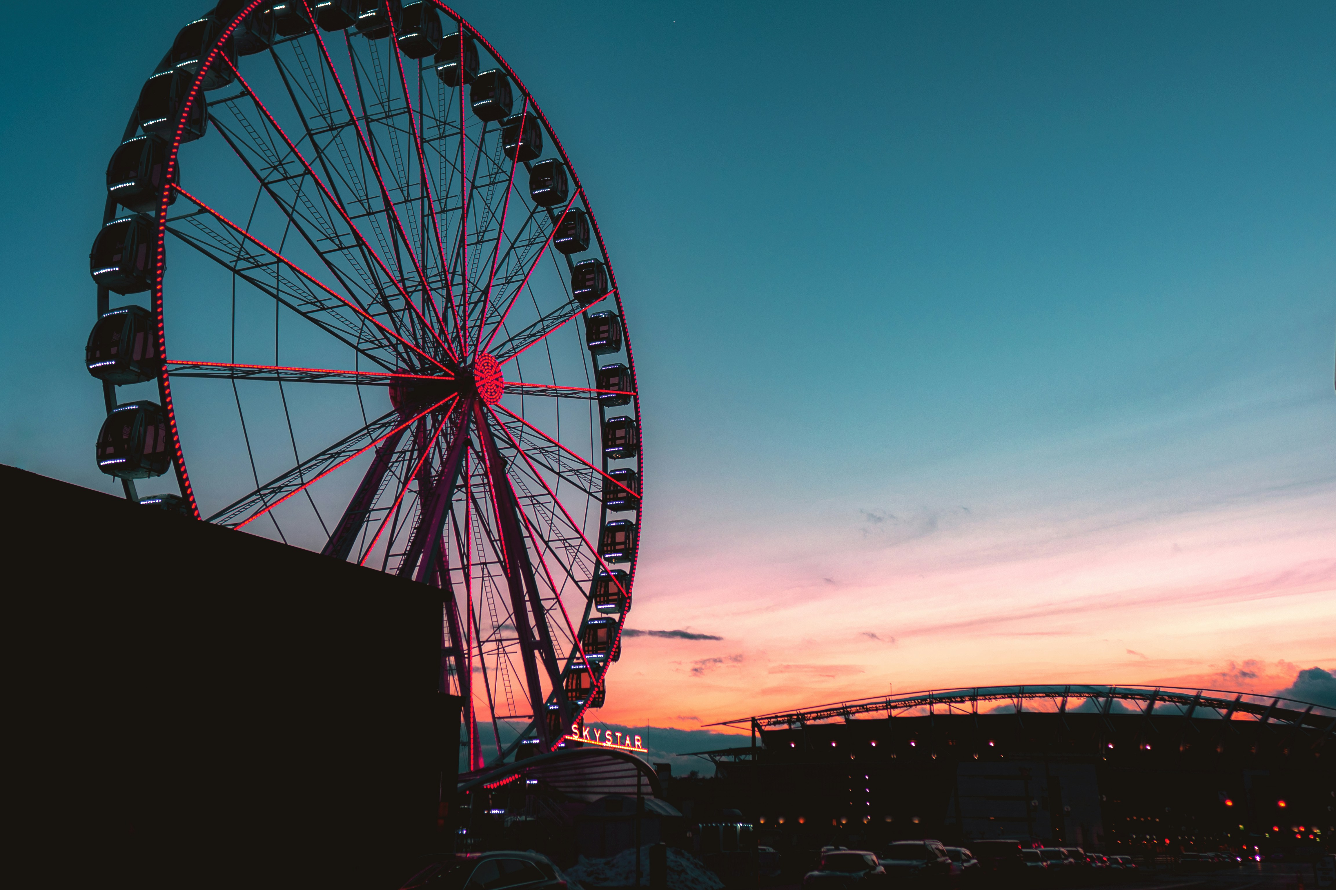 Ferris wheel illuminated at dusk with stadium silhouette against a vibrant sunset sky.