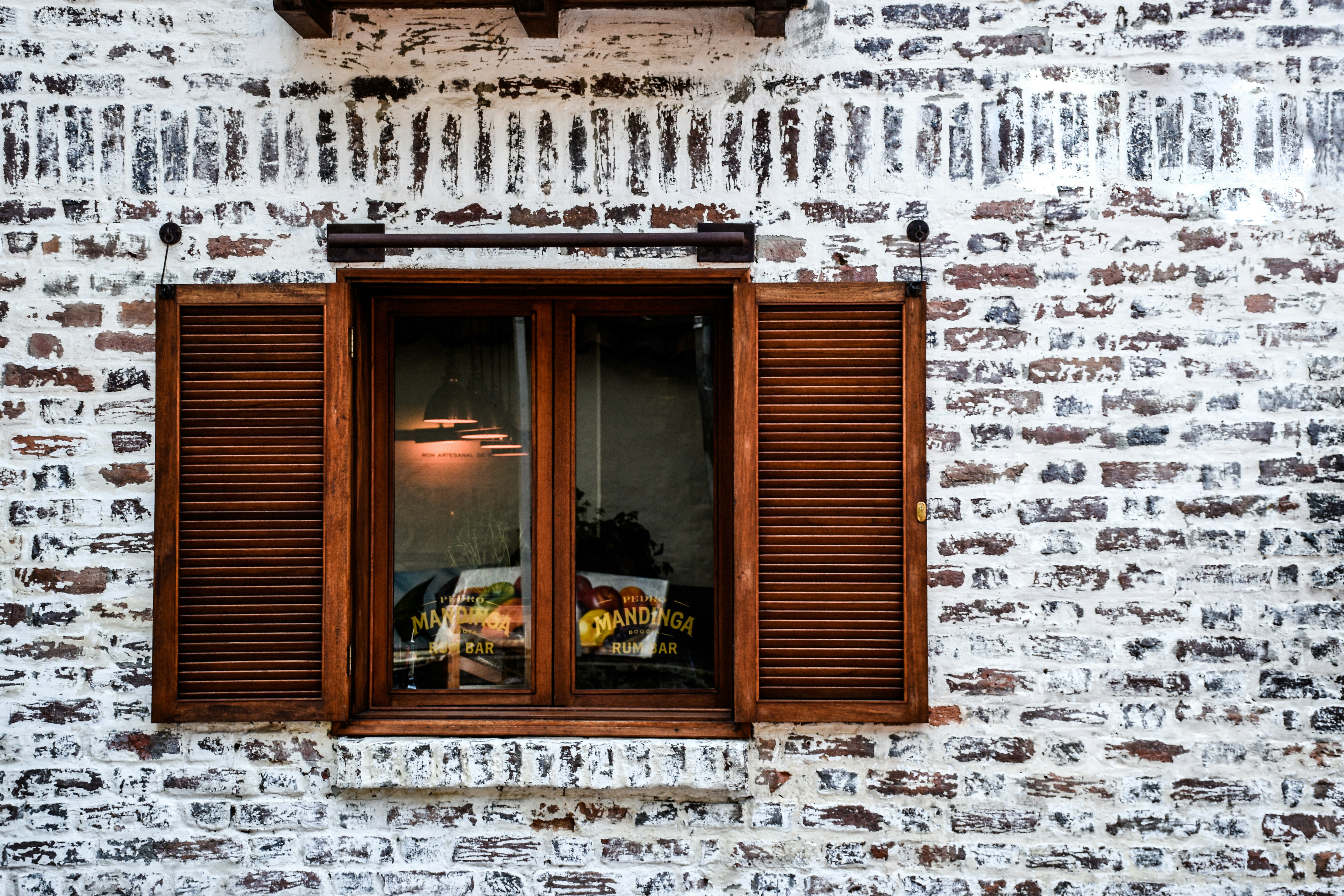 Wooden shutters frame a window revealing a softly lit interior, set against a textured white brick wall. The scene evokes a sense of nostalgia and warmth.