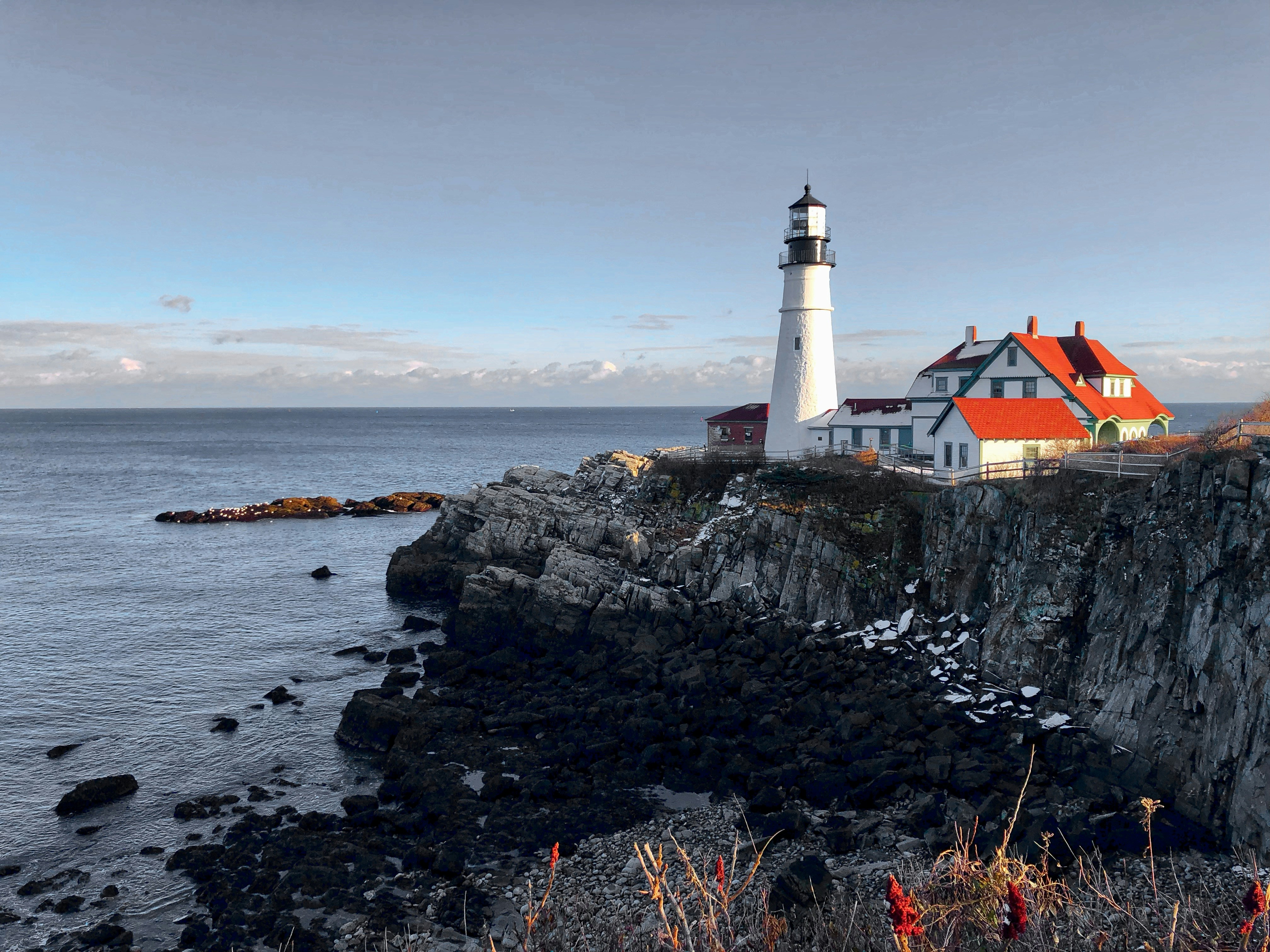 A historic lighthouse stands proudly on rugged cliffs overlooking the serene ocean, with a charming red-roofed house nearby. The scene captures the essence of coastal tranquility.