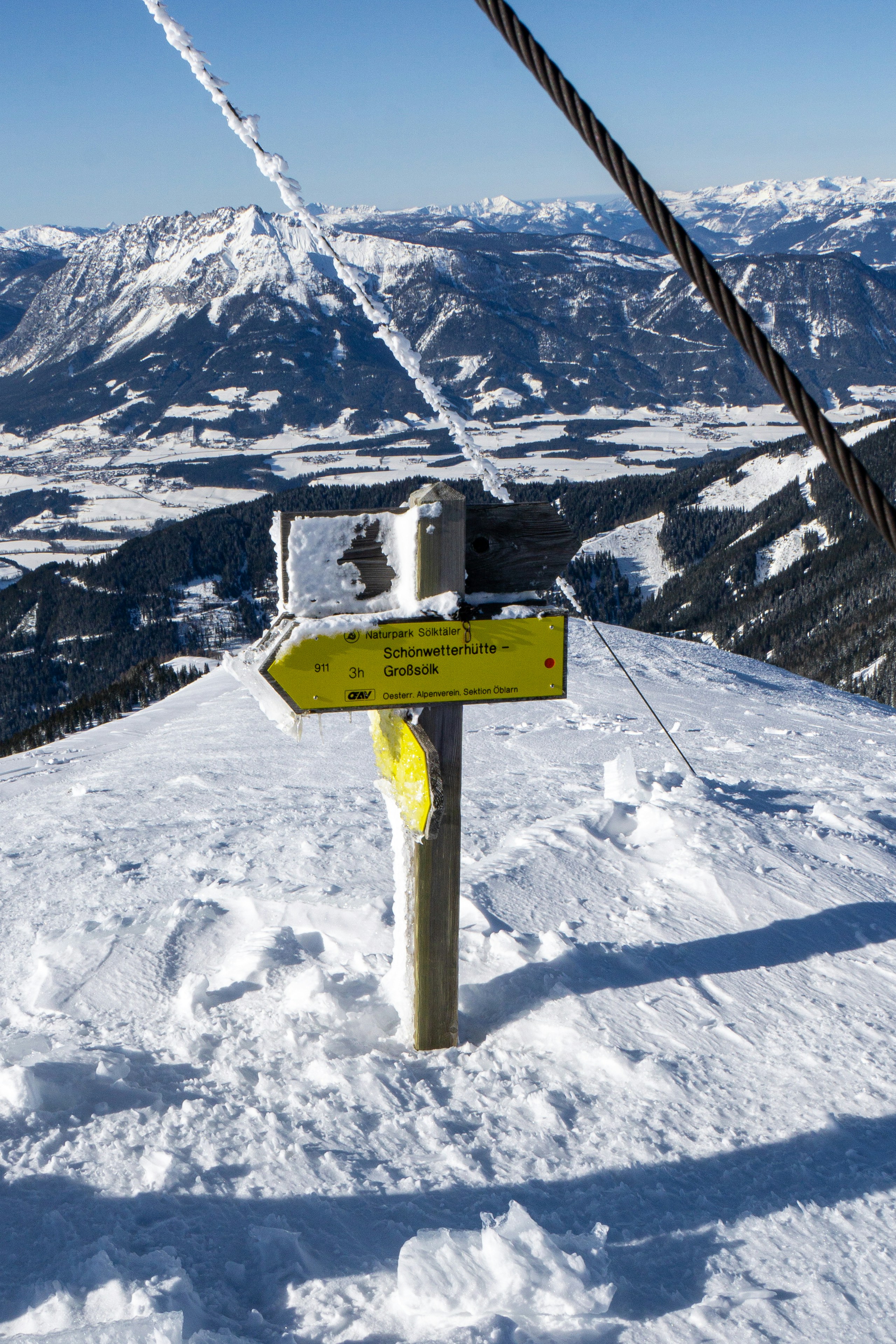 Snow-covered directional sign atop a mountain peak, guiding adventurers towards the Schönwetterhütte. The expansive view of the surrounding snow-capped mountains enhances the sense of adventure.