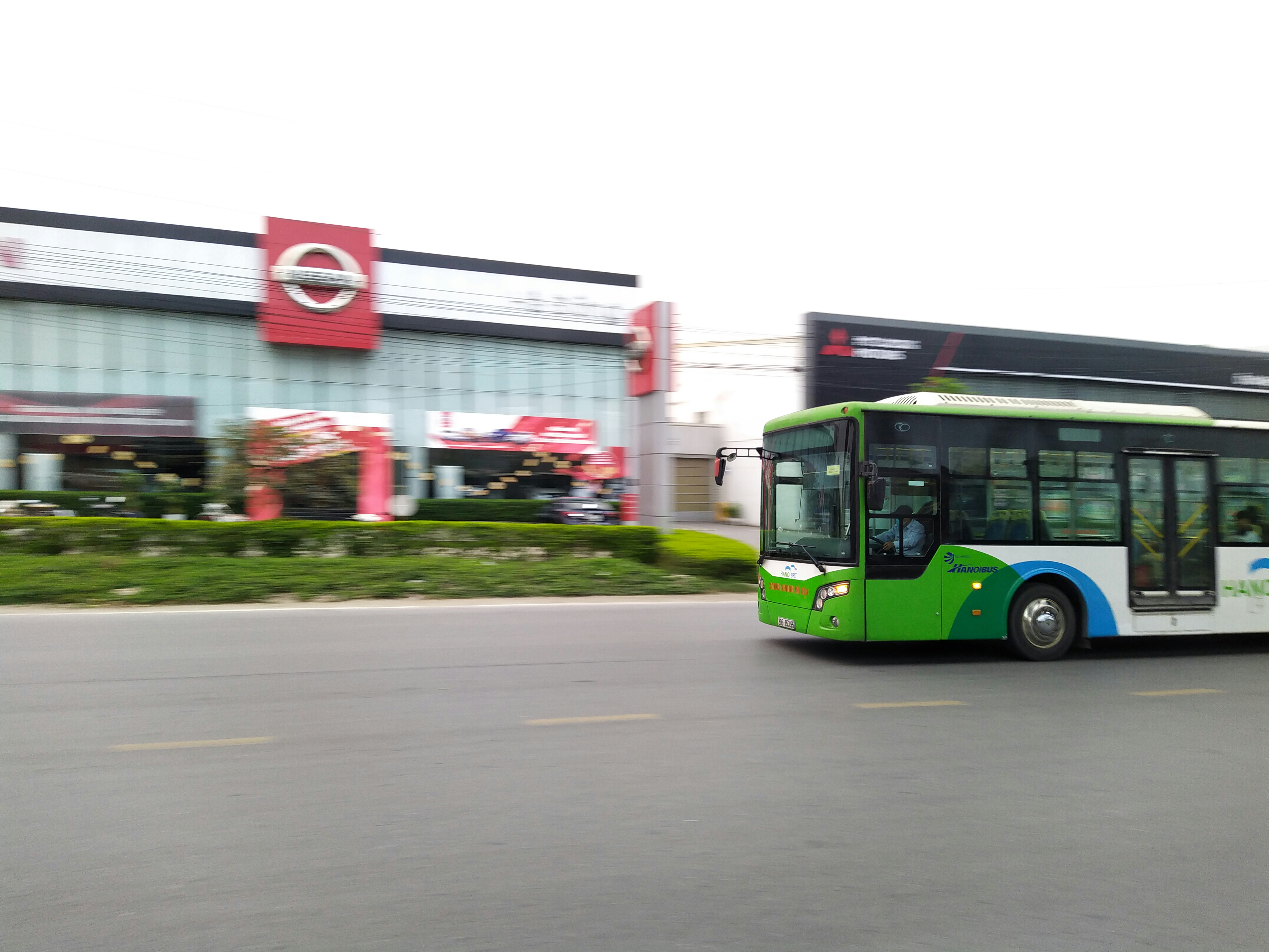Green and white city bus speeds past on a wide street, creating noticeable motion blur. A modern showroom with bold signage and angular architecture forms the backdrop.