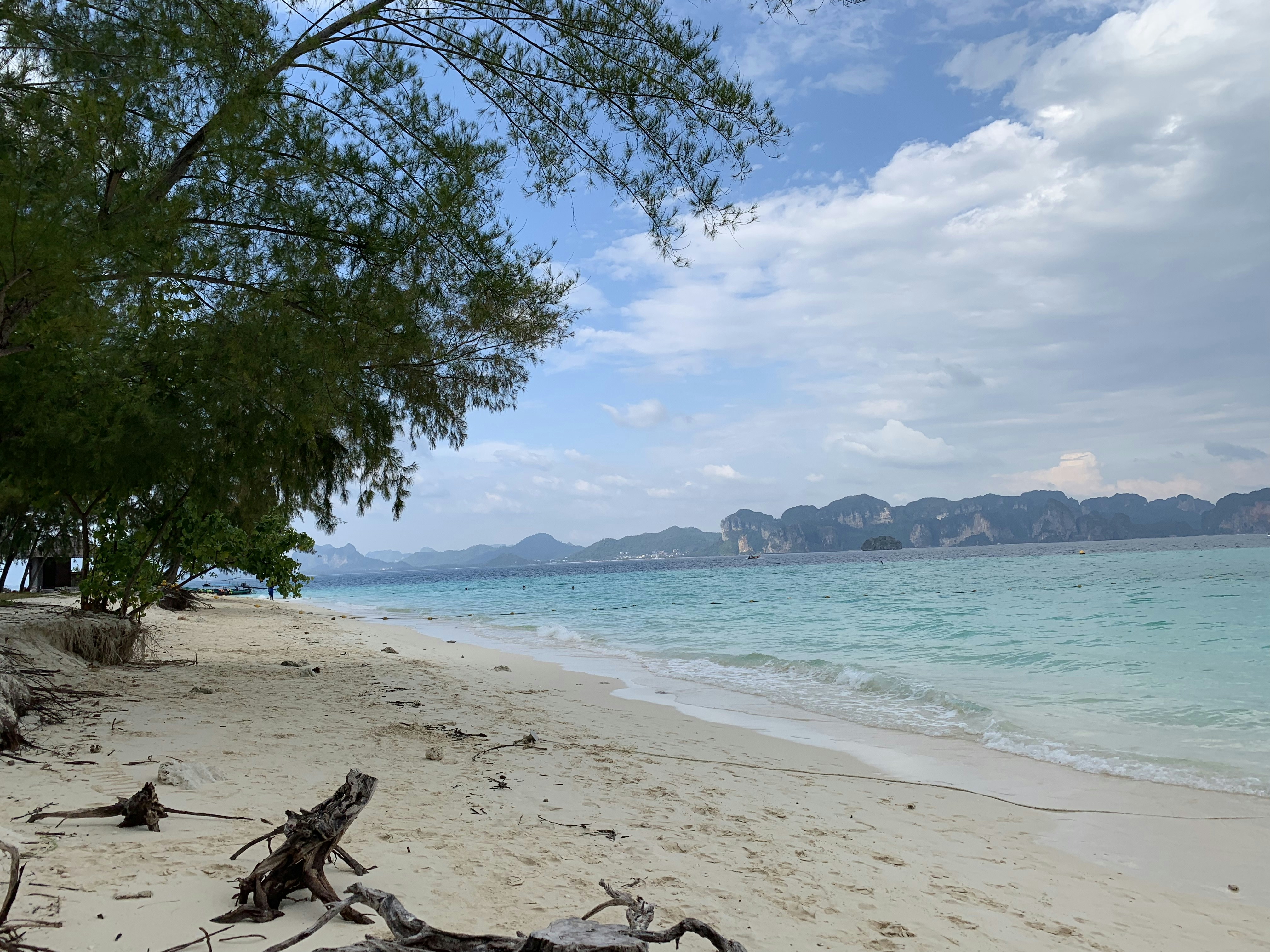 Serene beach scene featuring soft white sand, gentle waves, and distant limestone cliffs under a partly cloudy sky.