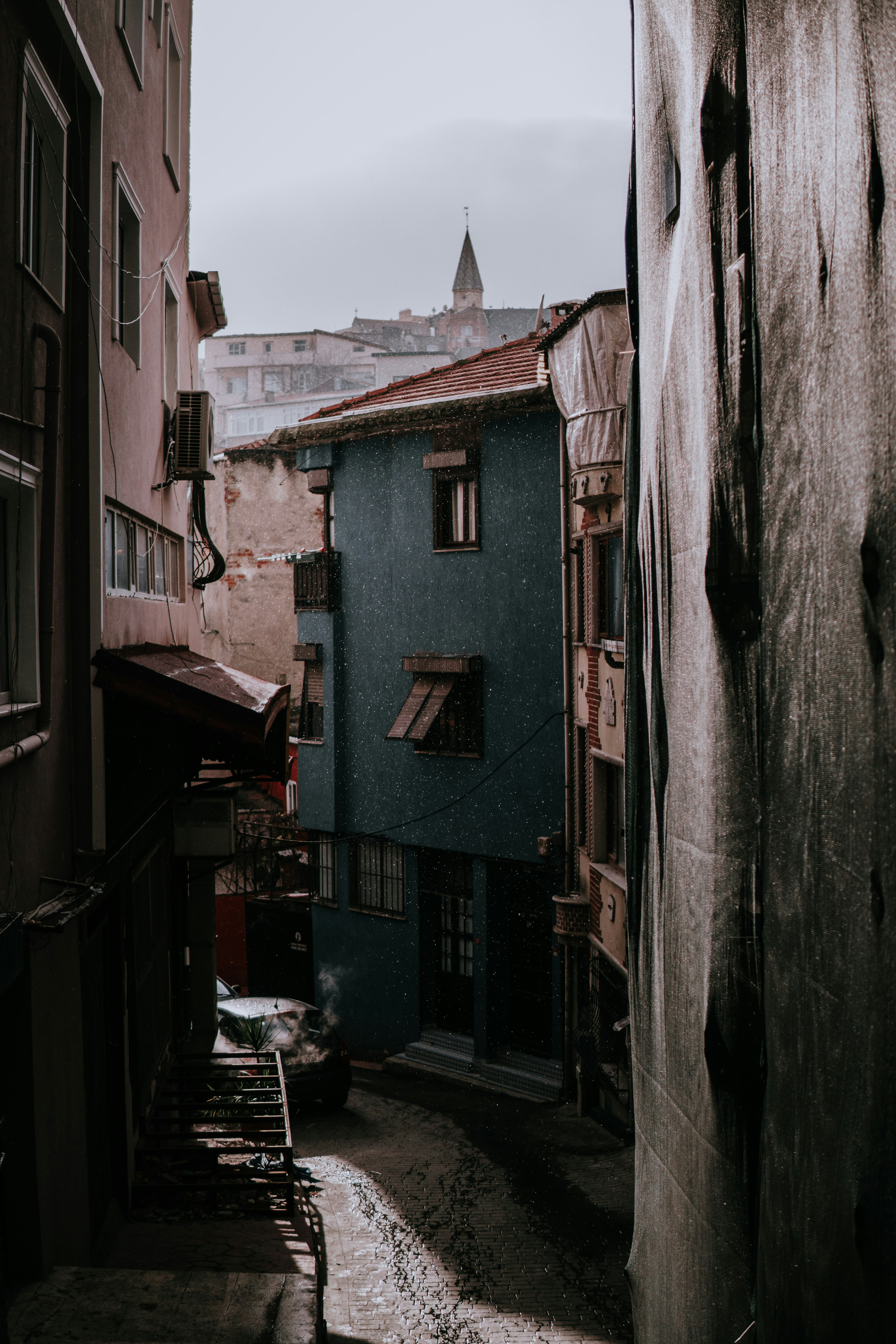 Narrow alleyway framed by towering buildings, showcasing a striking blue facade and distant architectural details. Light filters through, creating a moody atmosphere.