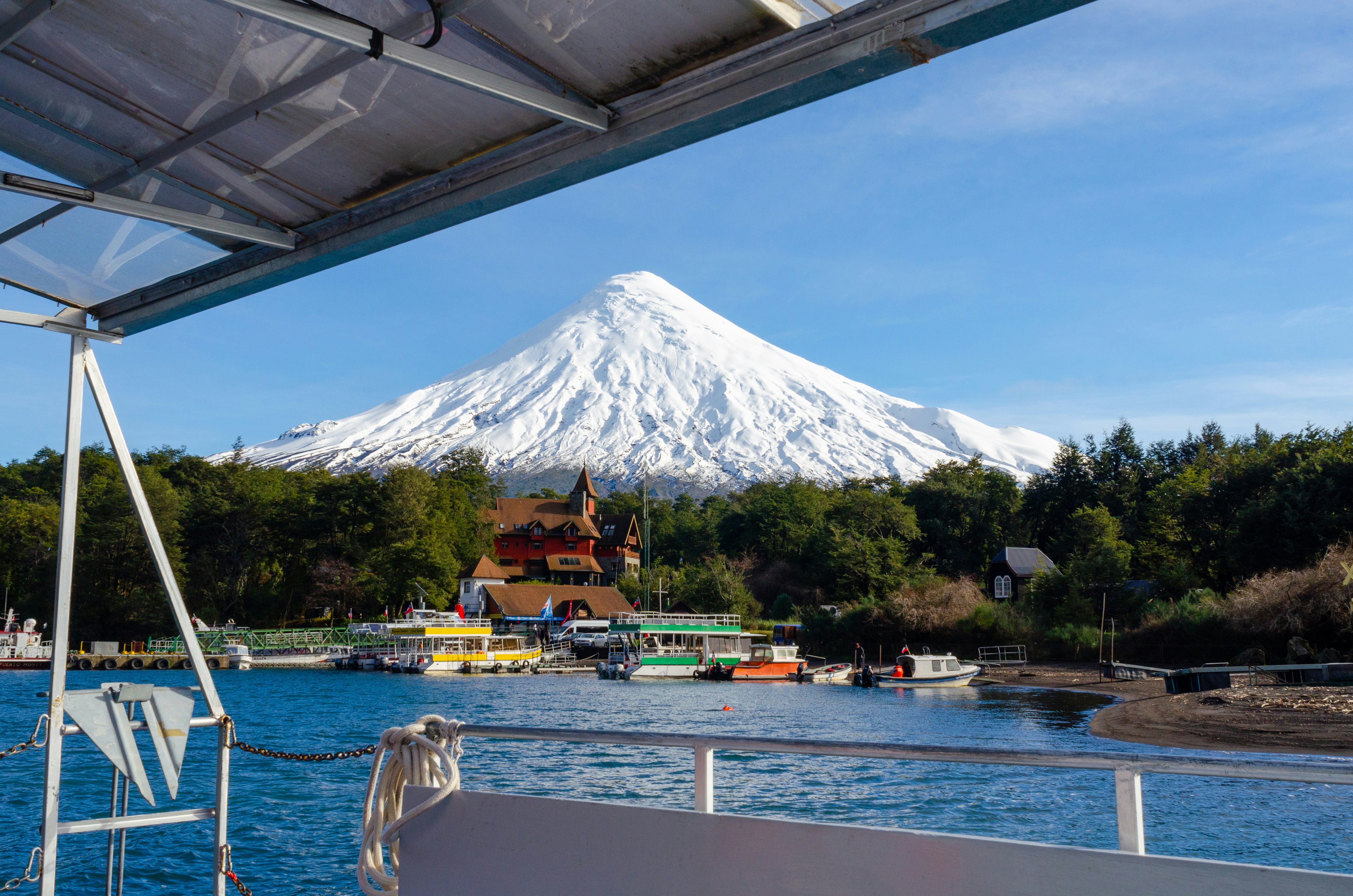 Snow-capped mountain towers over a tranquil harbor, framed by lush greenery and colorful boats. A picturesque scene capturing nature's grandeur.