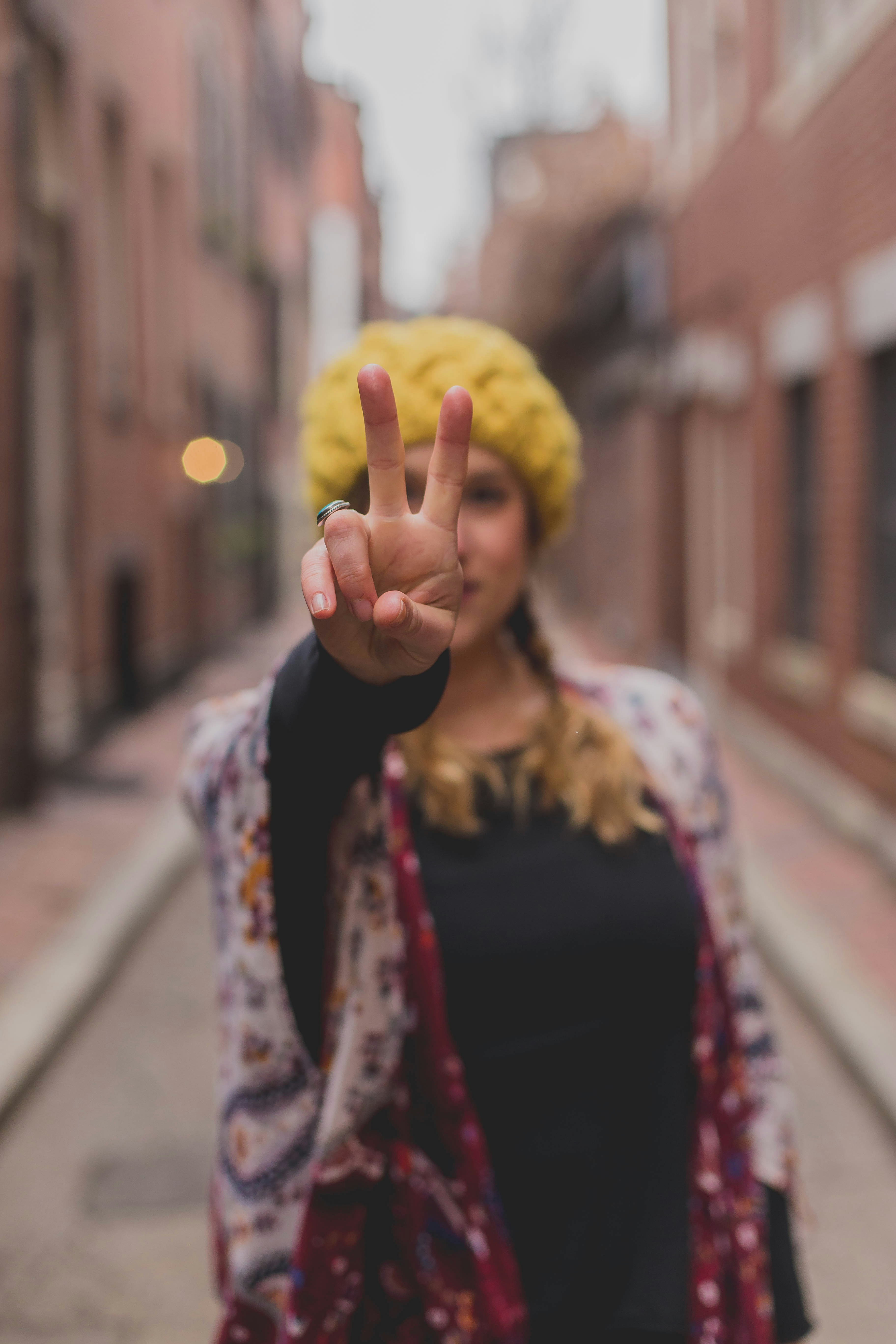 A woman in a vibrant yellow hat flashes a peace sign against a blurred alley backdrop. The urban setting enhances the sense of tranquility amidst city life.