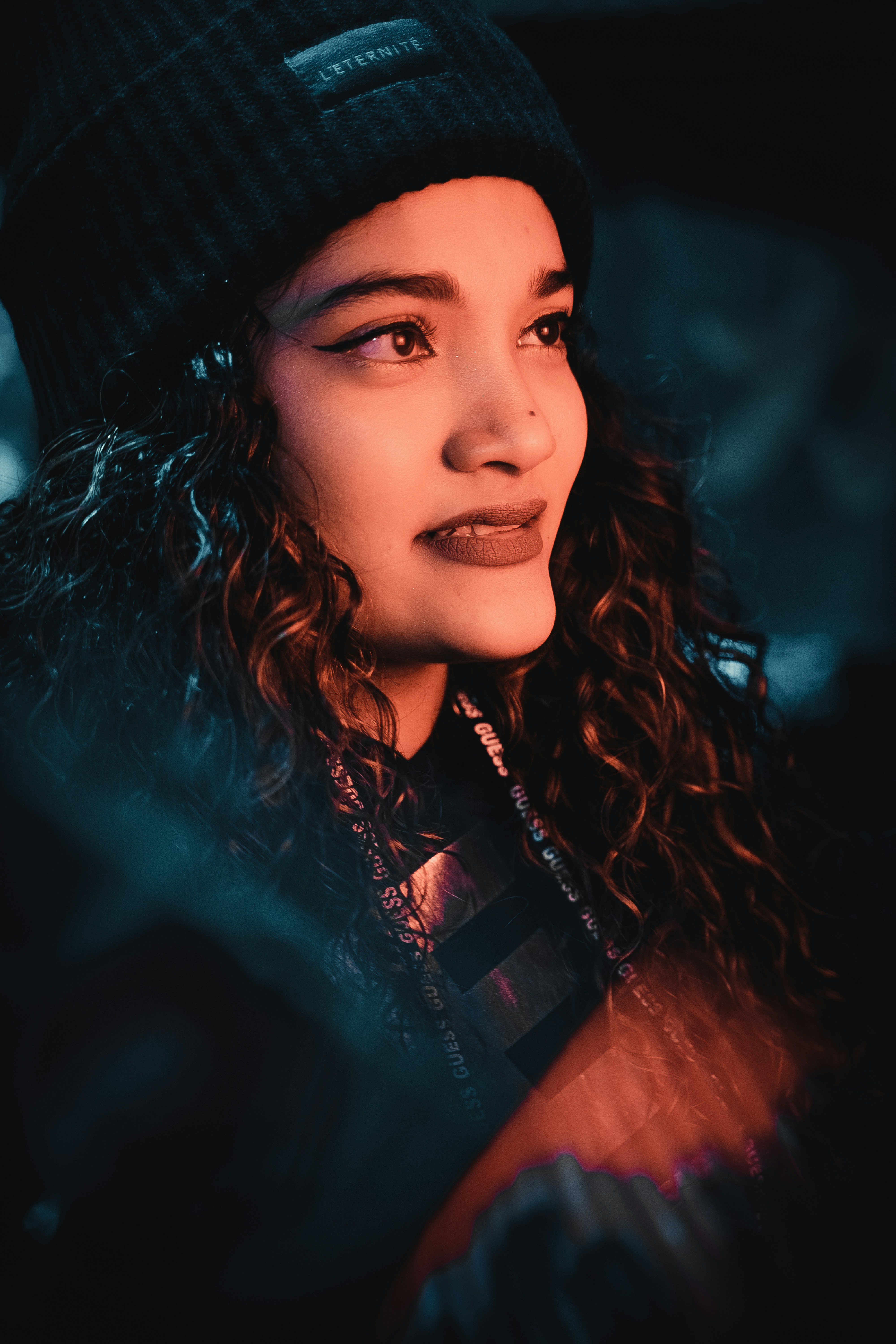 A young woman with curly hair and a beanie, illuminated by soft, colorful lighting, reflects a moment of contemplation in an urban setting.