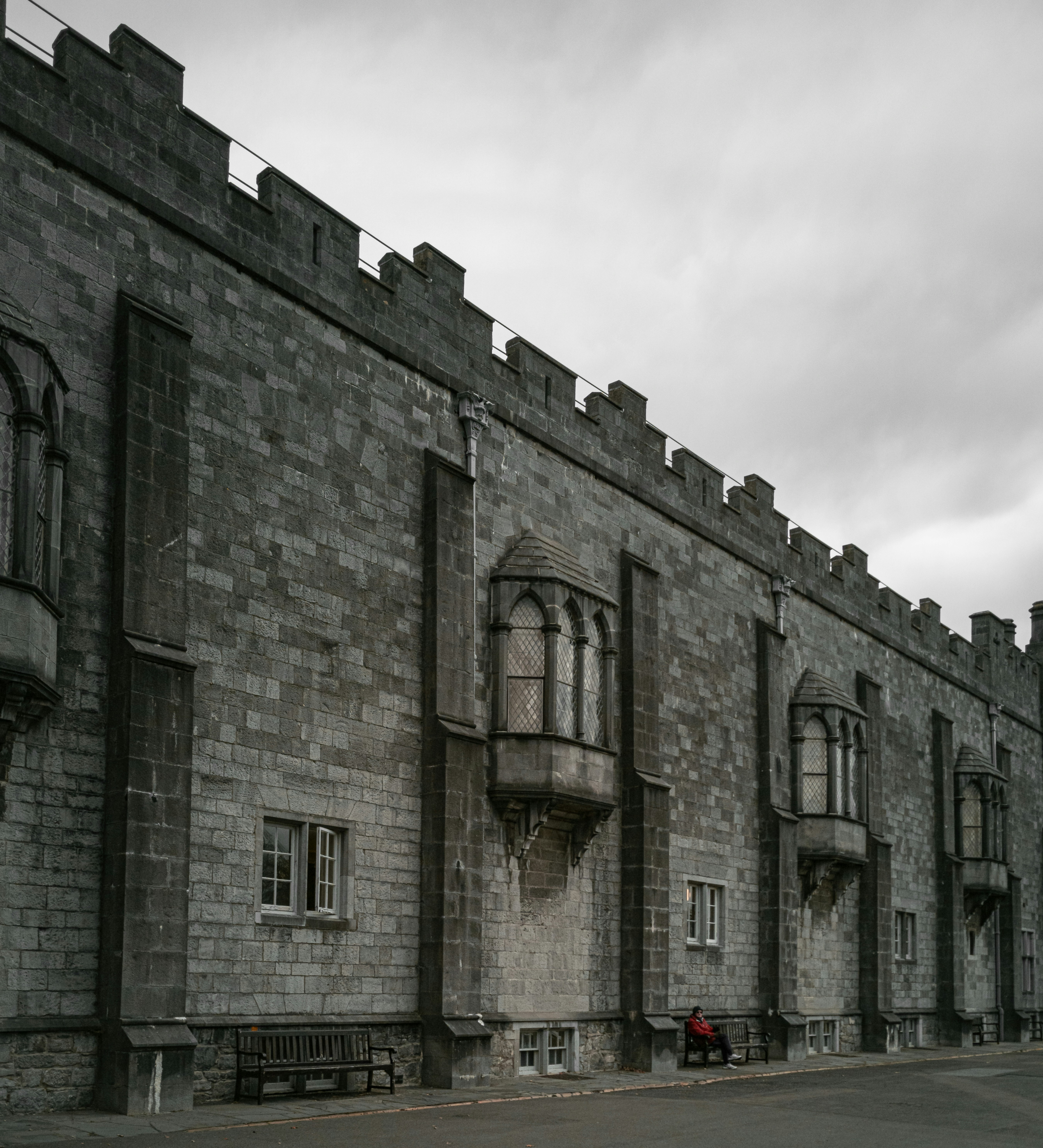 Historic stone wall adorned with ornate window structures, featuring a solitary figure seated nearby. 