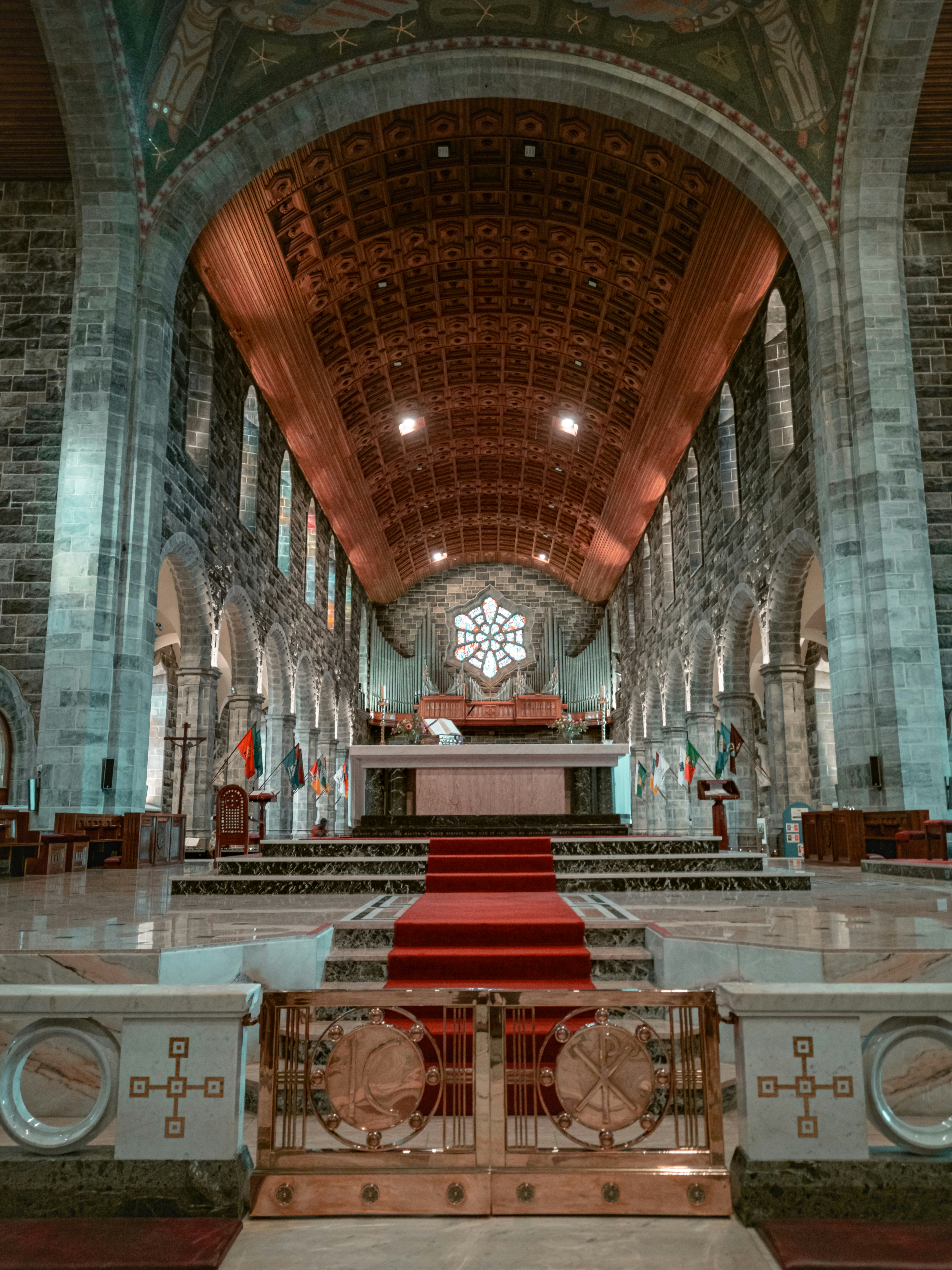 Interior of a grand church showcasing intricate stonework and a stunning wooden ceiling, highlighted by colorful stained glass. Red carpet leads to the altar.