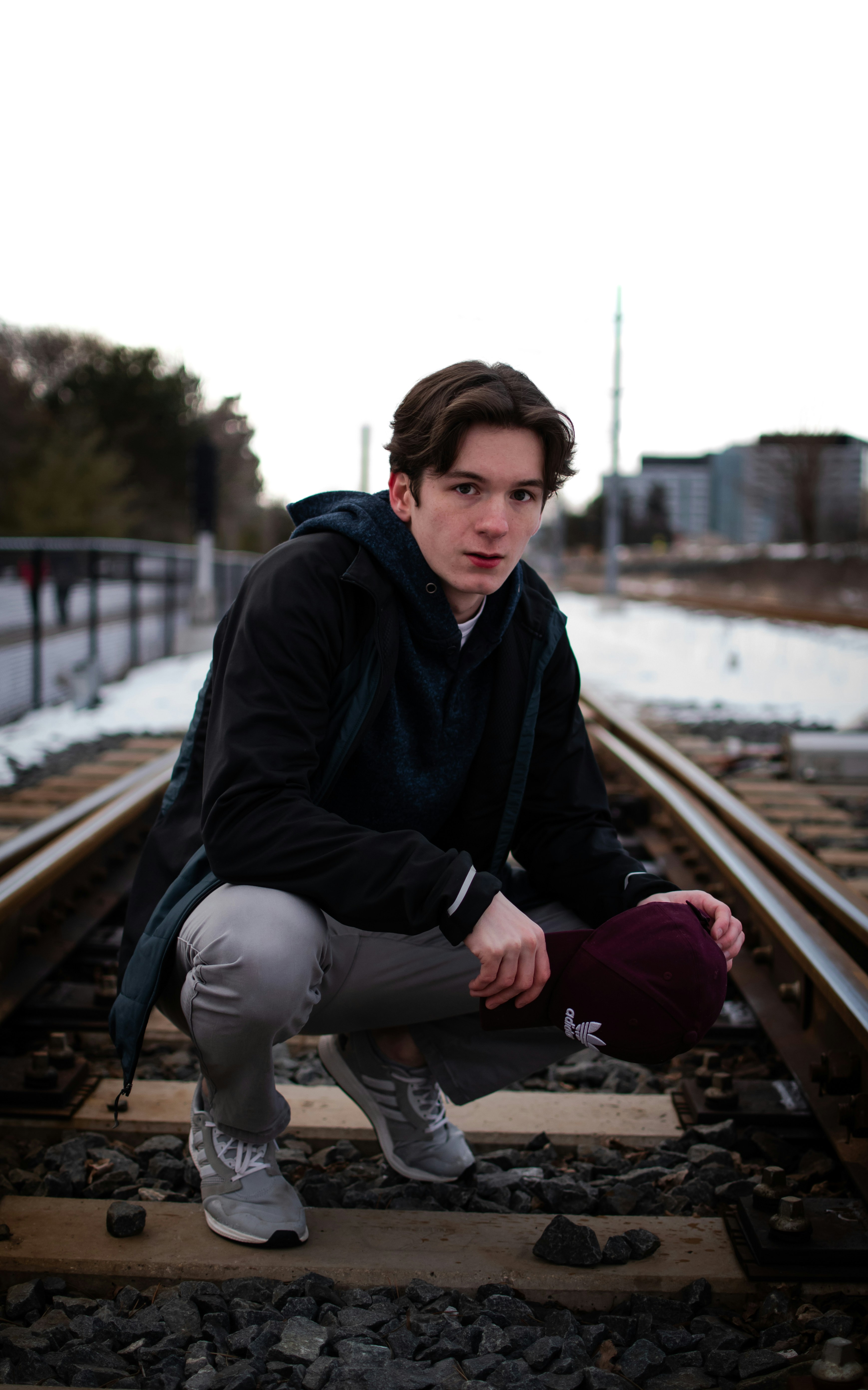 Portrait of a man sitting on railway tracks holding a red adidas hat on a cold winter evening with winter jacket and hoodie