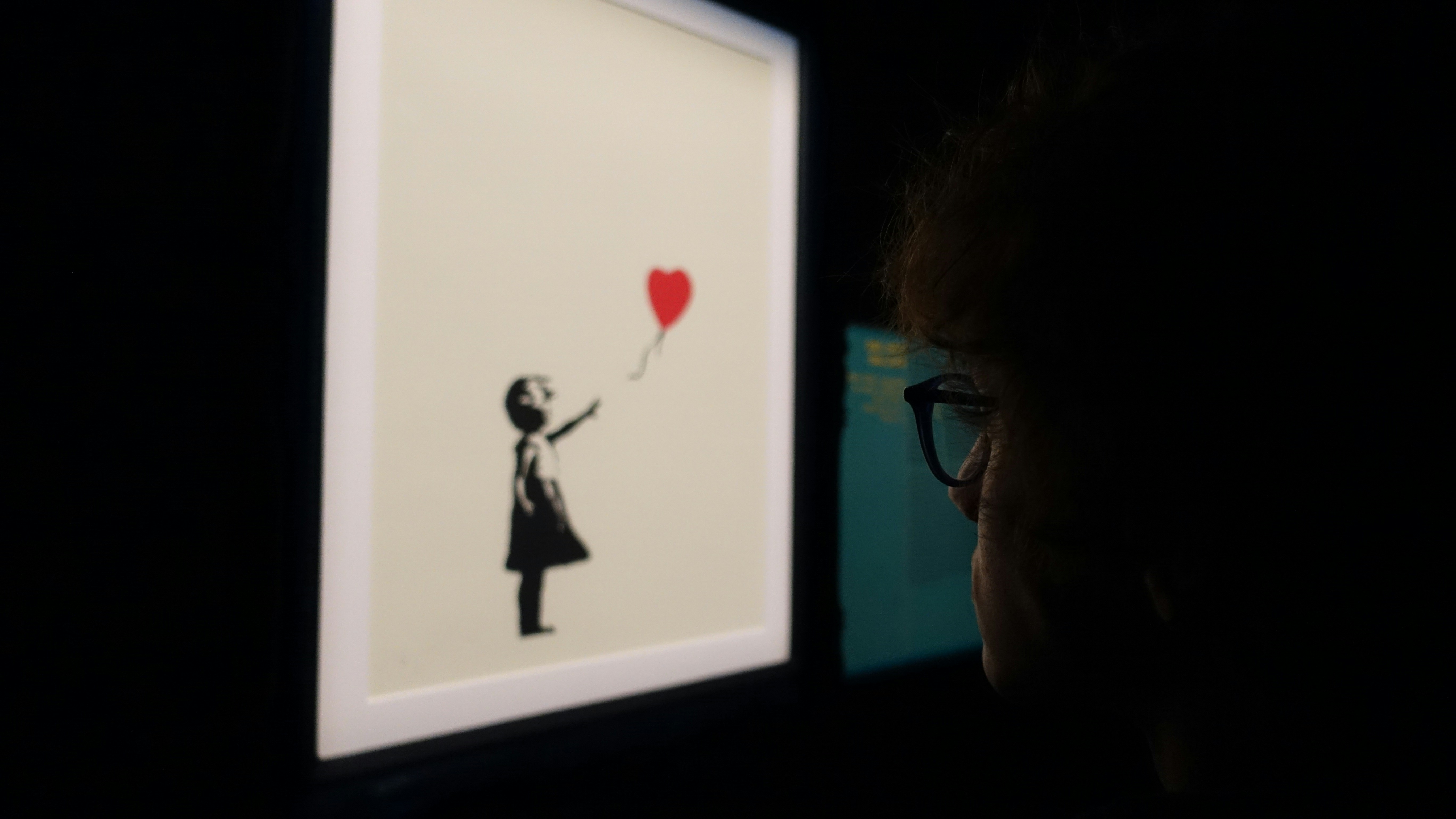 Silhouette of a person observing artwork of a girl reaching for a red heart-shaped balloon.
