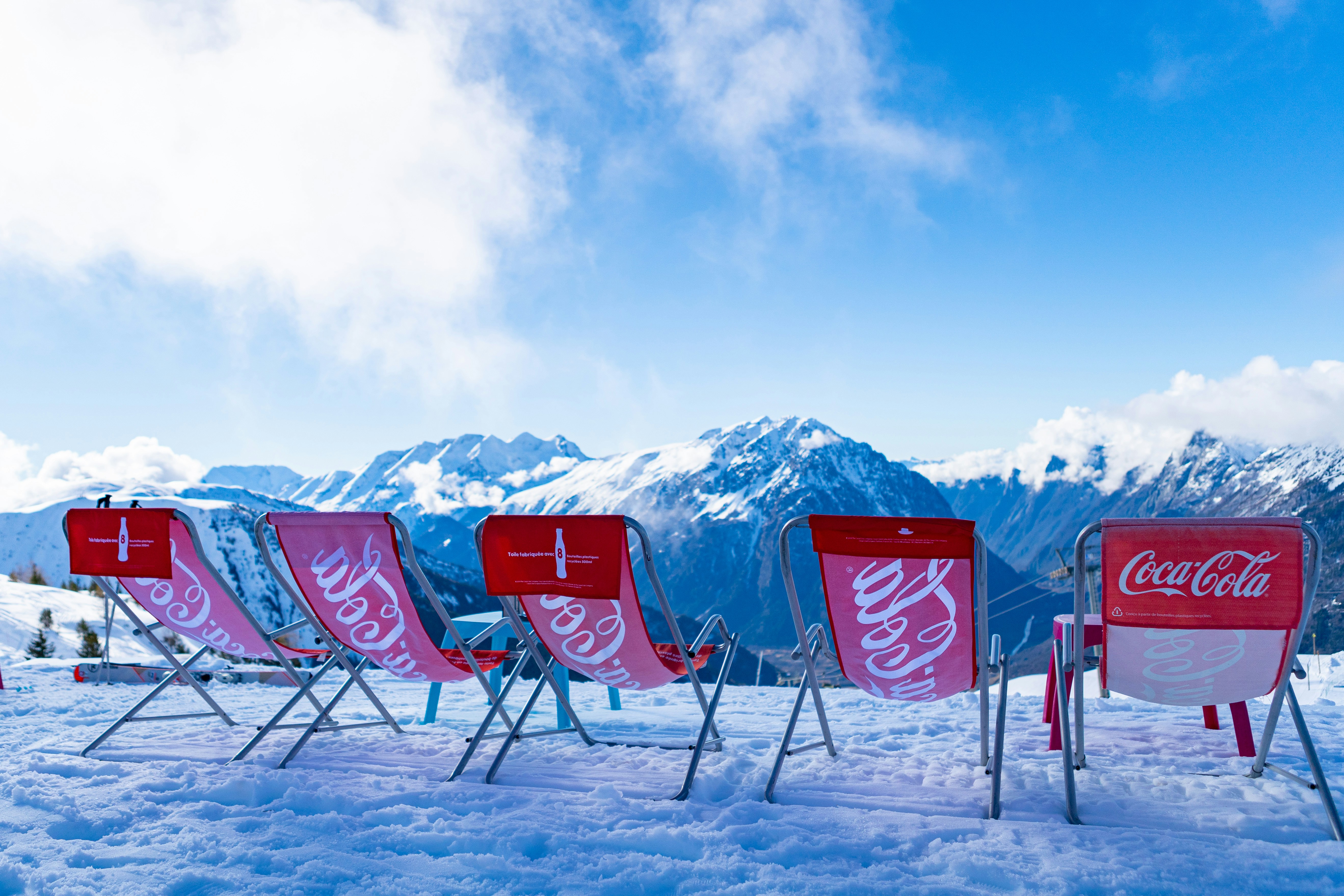 Five red lounge chairs set against a backdrop of snow-covered mountains, inviting relaxation under a clear blue sky.