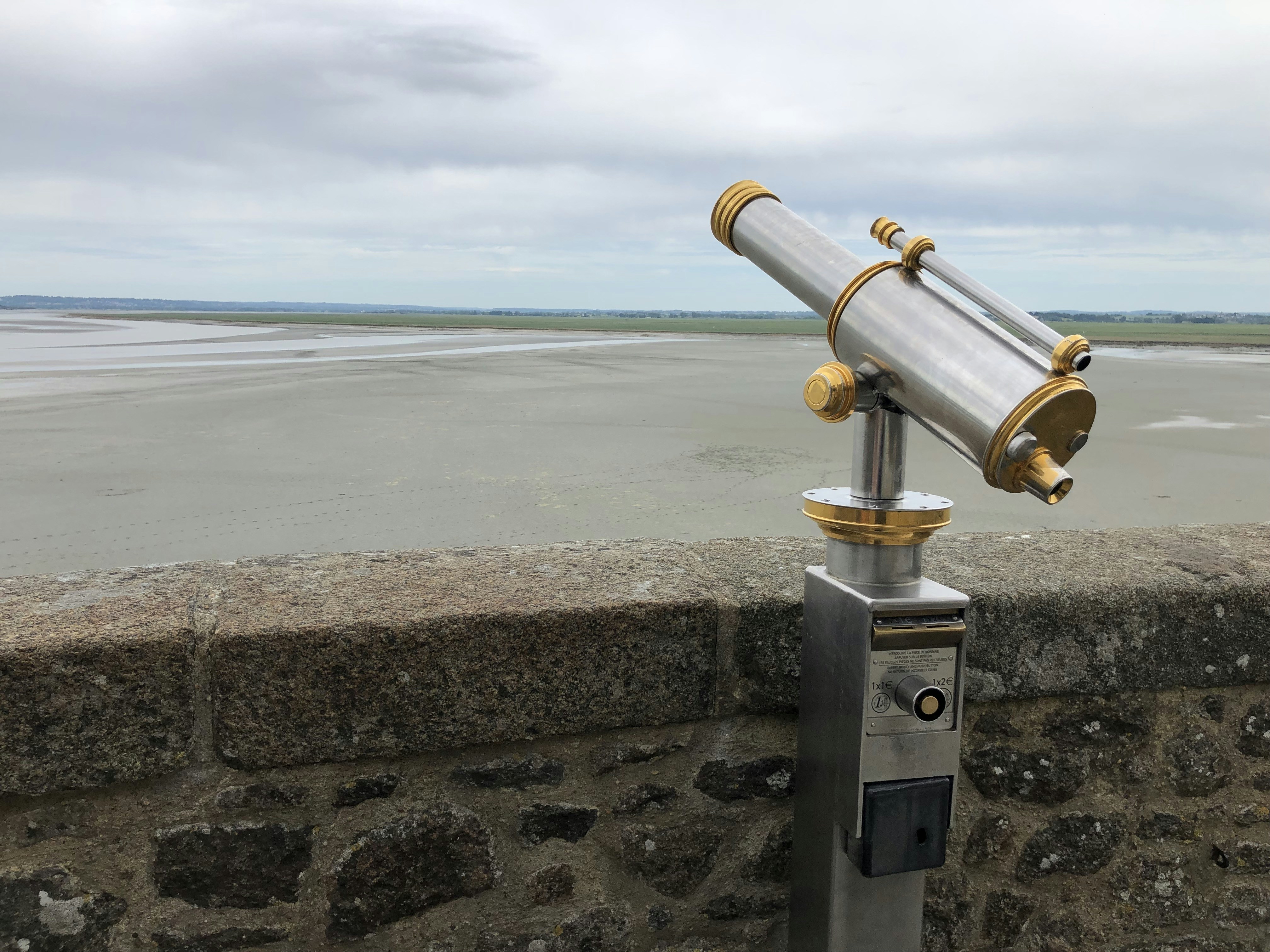 A metallic telescope mounted on a stone wall overlooking a vast, empty coastline under a cloudy sky.