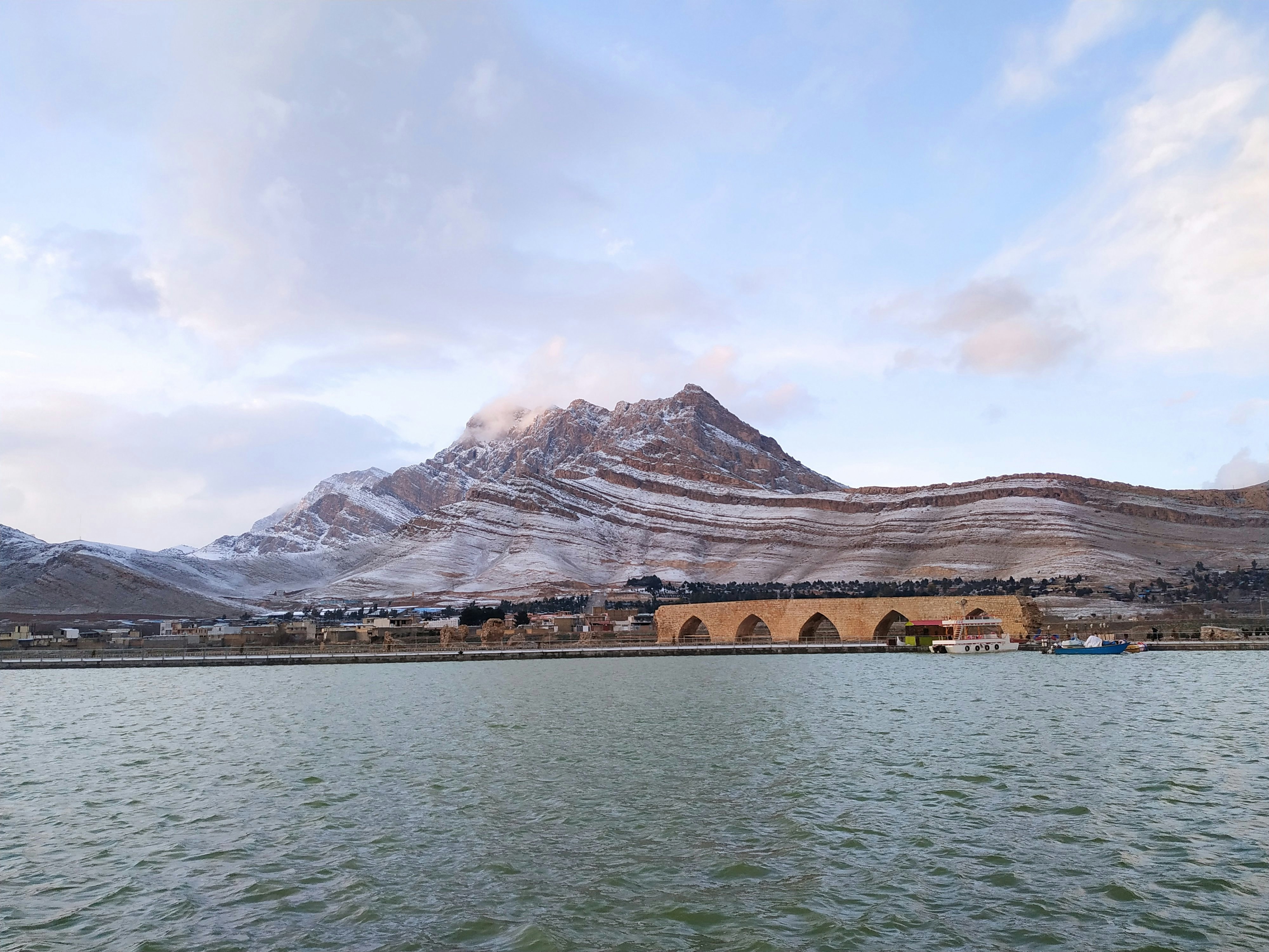 Mountain with snow and a historic bridge over a tranquil lake under a partly cloudy sky.