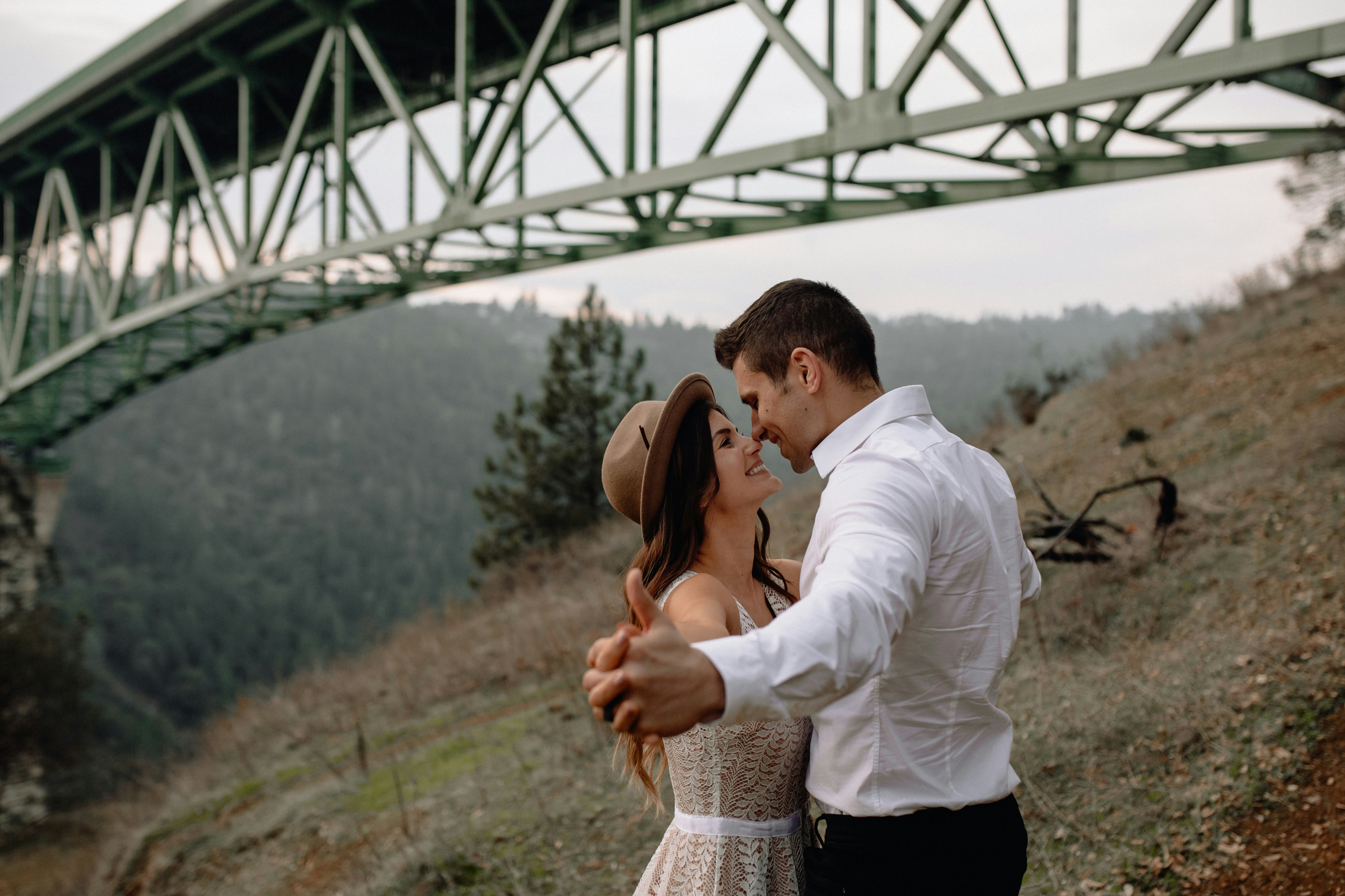 A couple shares an intimate moment beneath a towering green bridge, surrounded by lush greenery and rolling hills. The soft, natural light casts a gentle glow on their faces, highlighting the contrast between the rustic landscape and their elegant attire. The image captures a serene yet passionate atmosphere, with the geometric lines of the bridge adding a striking architectural element to the composition.