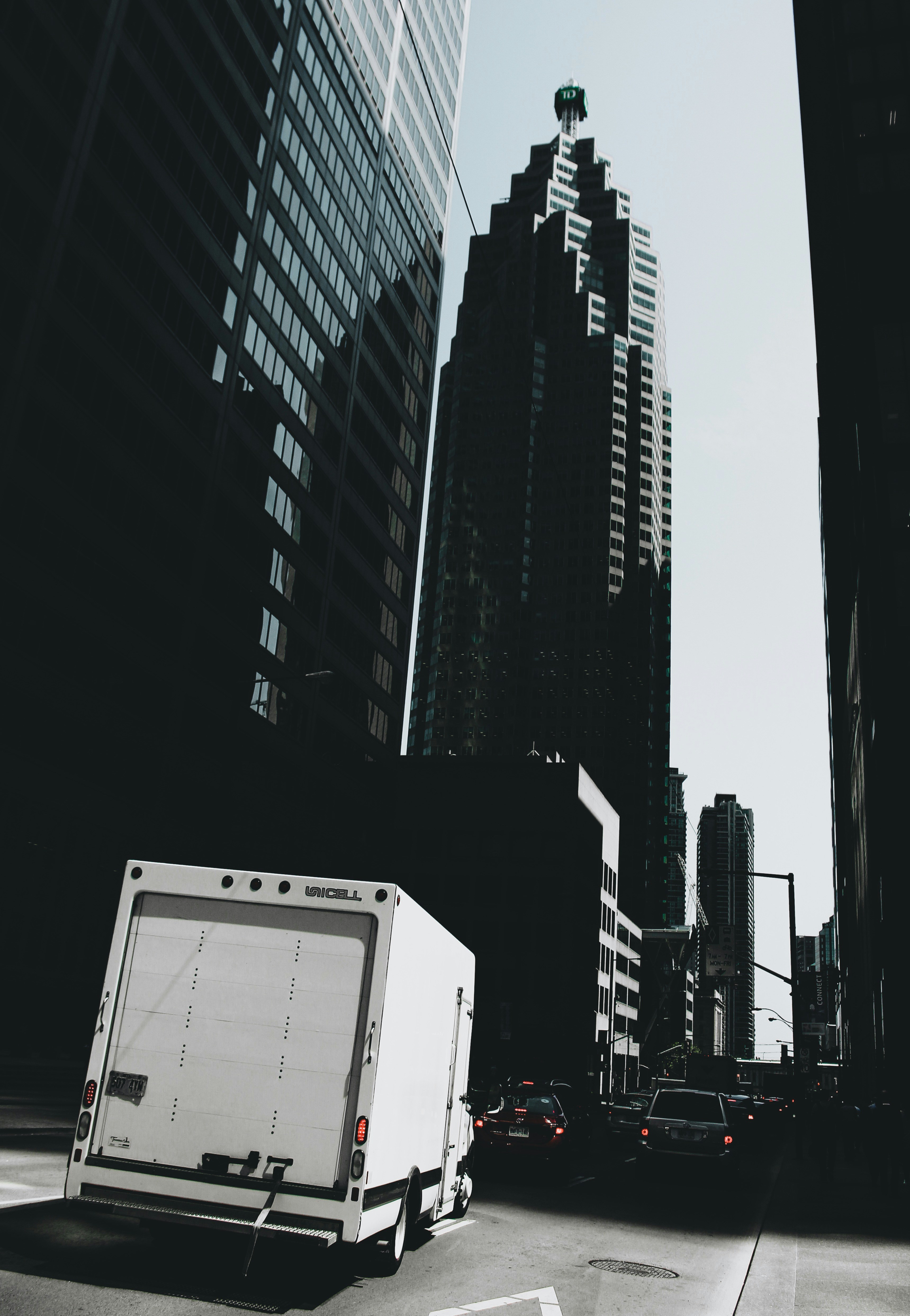 A white delivery truck navigates through a bustling cityscape, flanked by towering skyscrapers under a bright sky.