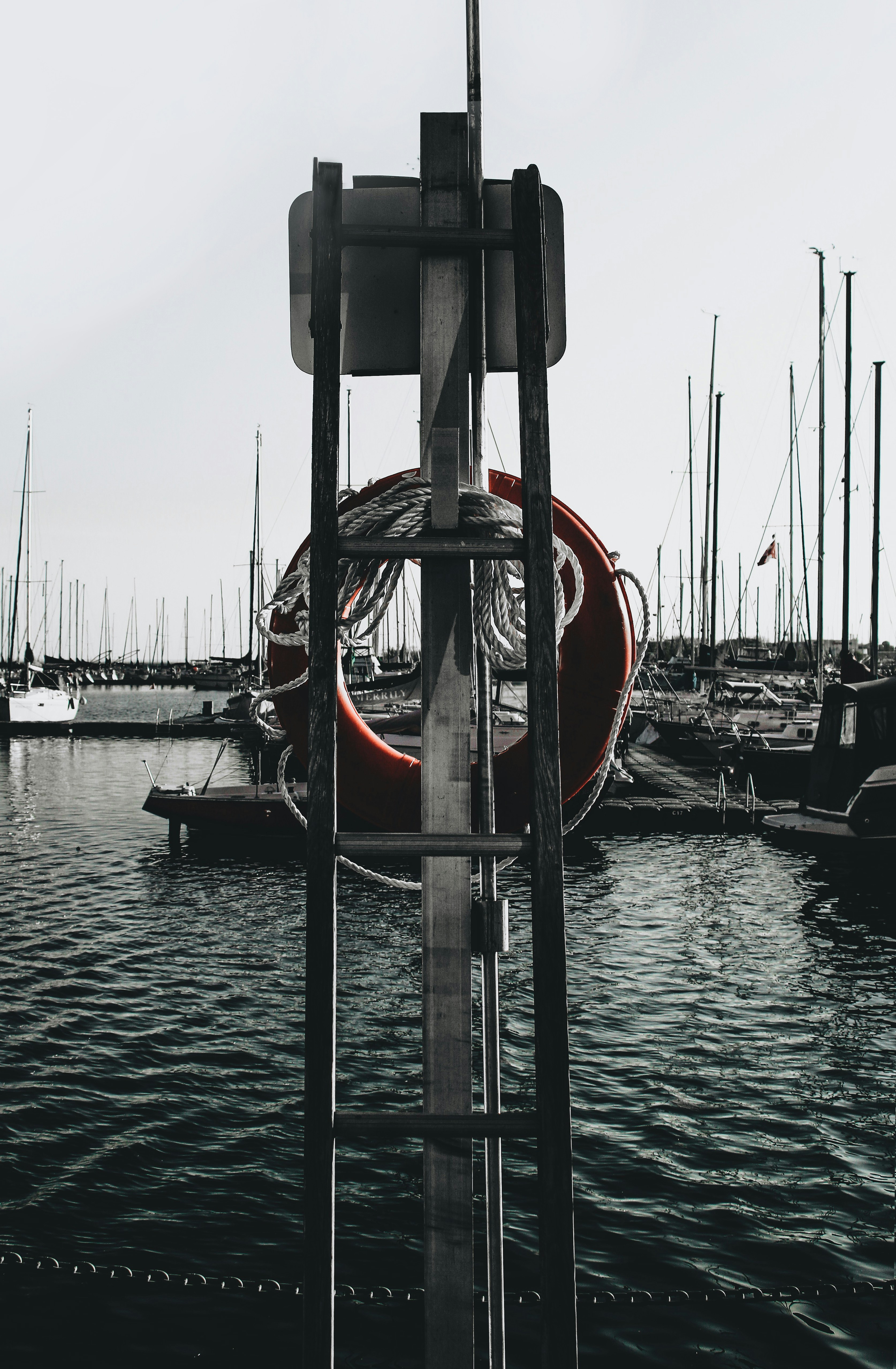 A life ring suspended on a wooden frame, overlooking a tranquil harbor filled with sailboats. The scene captures the essence of maritime safety against a backdrop of calm waters.
