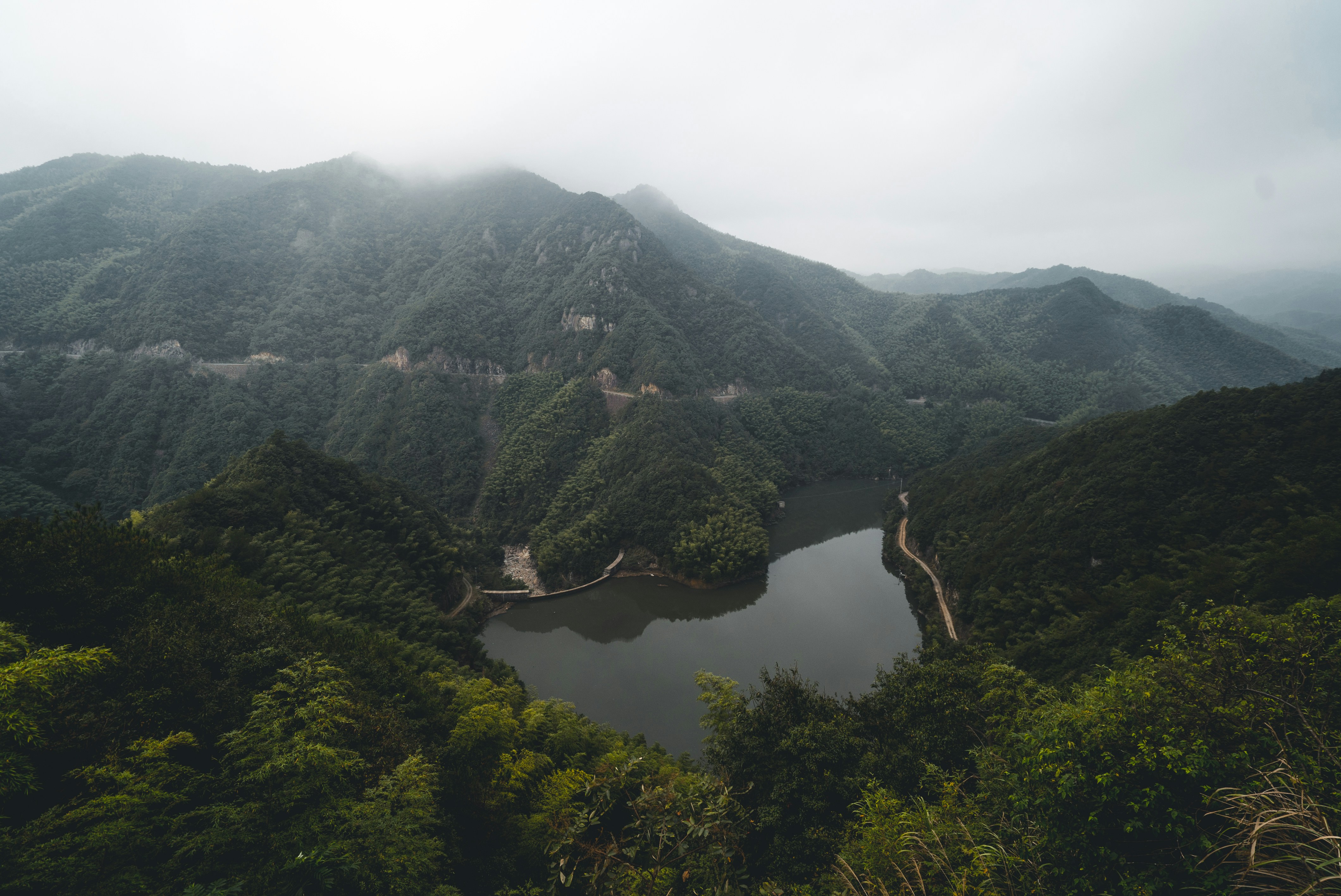 Misty mountains envelop a tranquil lake with lush greenery and a winding road.