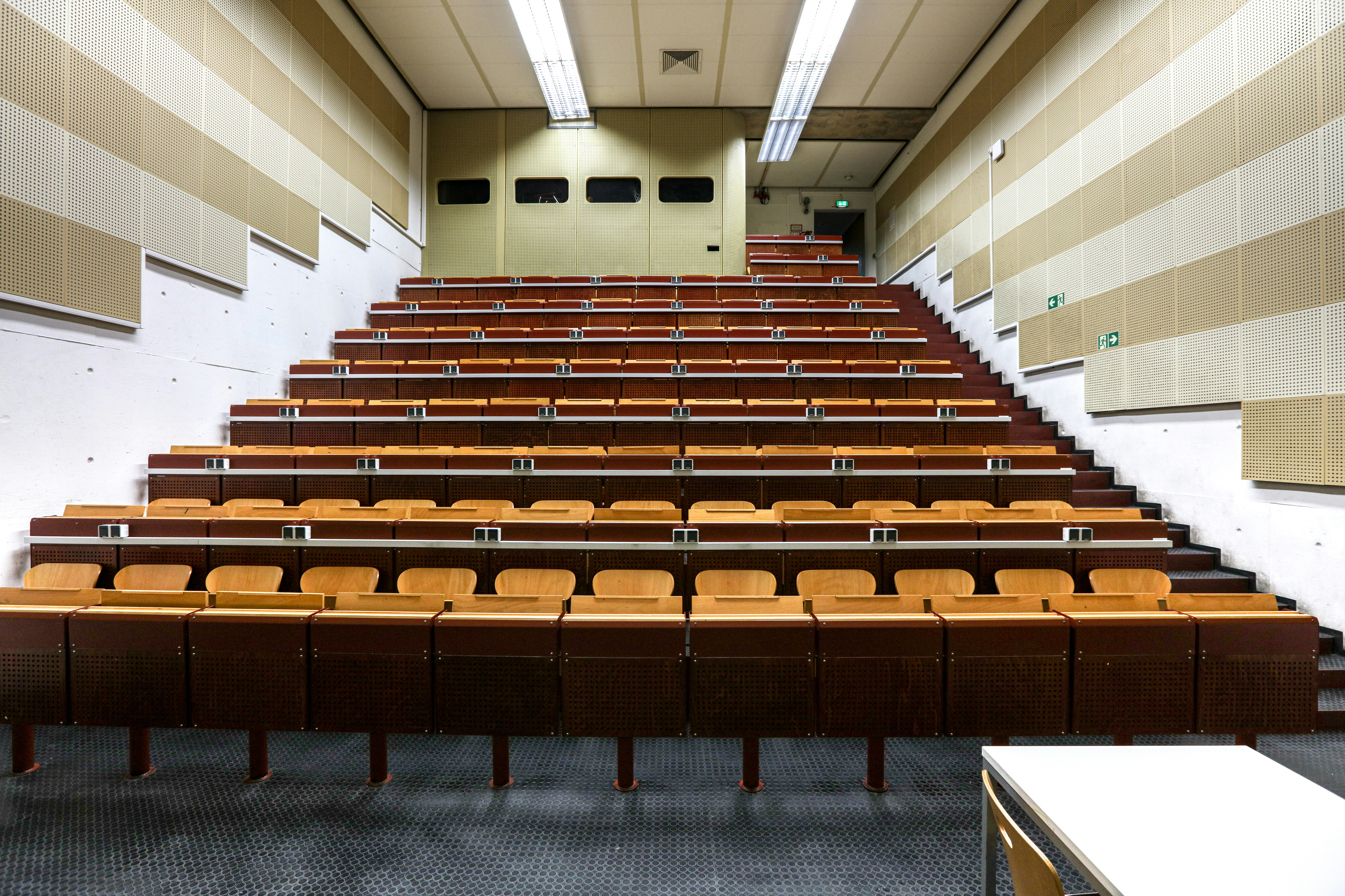 Empty lecture hall with rows of wooden seats facing a whiteboard.