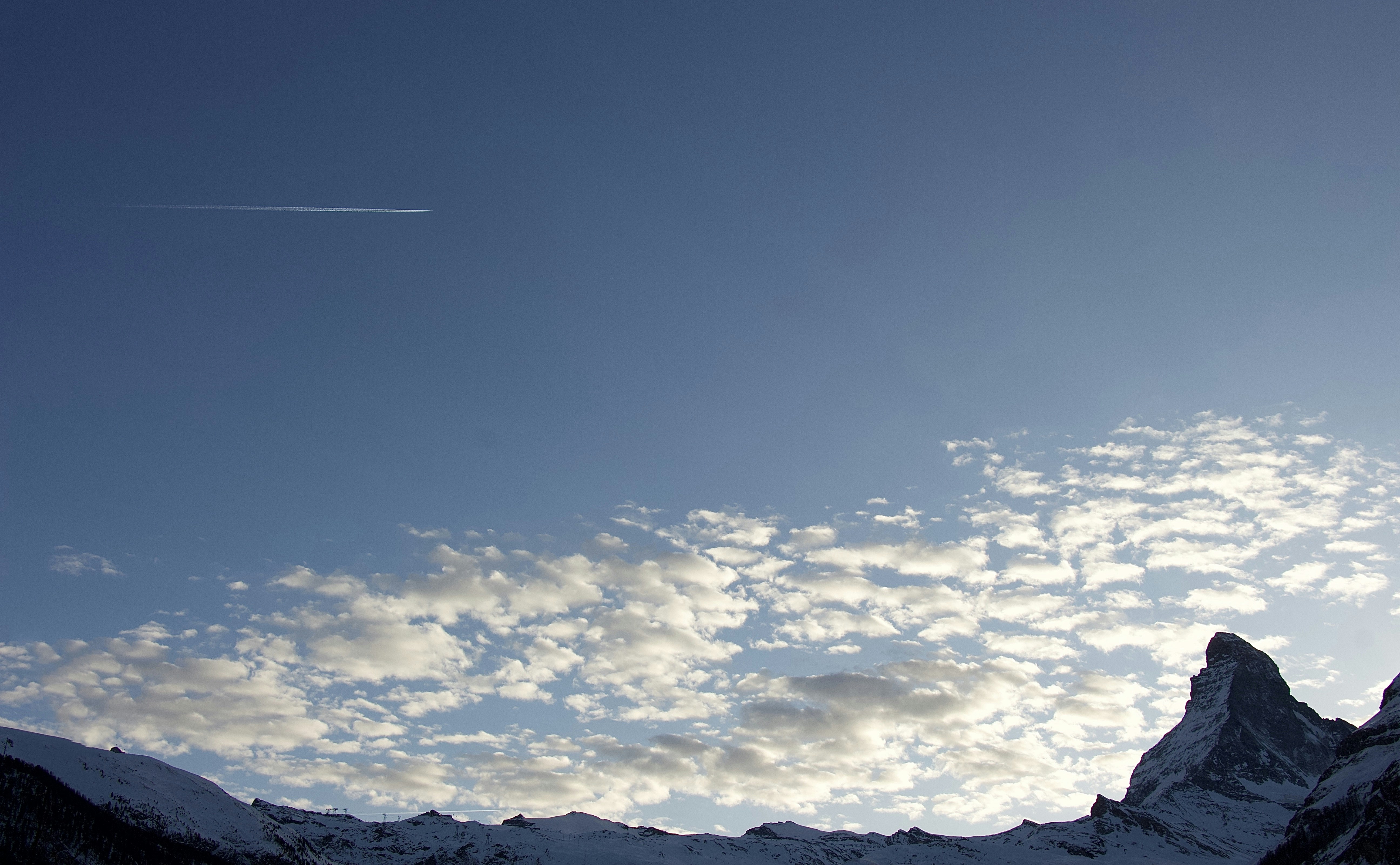 Snow-capped mountain under a sky dotted with scattered clouds and a distant contrail.