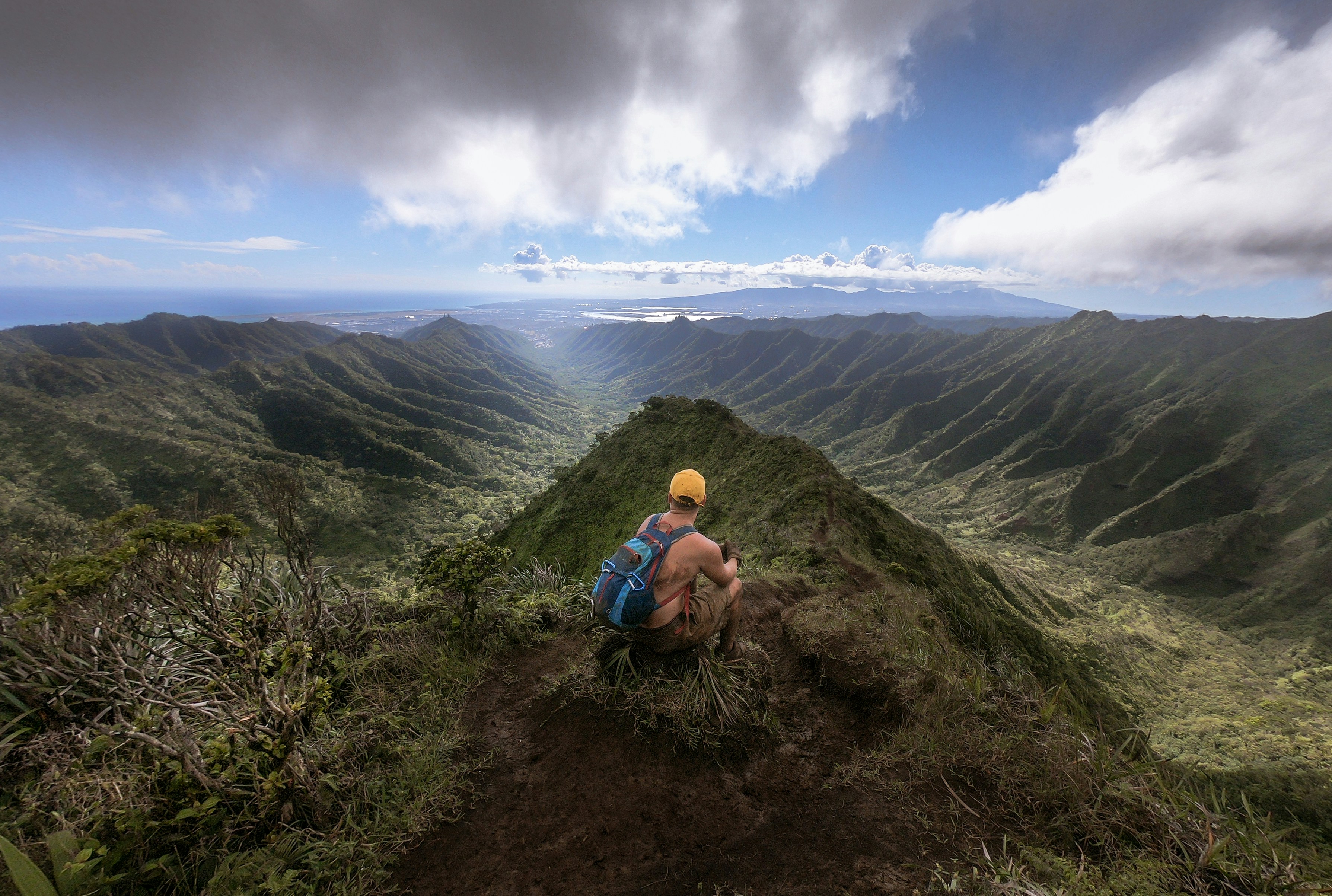 Hiker perched on a narrow ridge overlooking lush Hawaiian valleys under a dramatic sky.