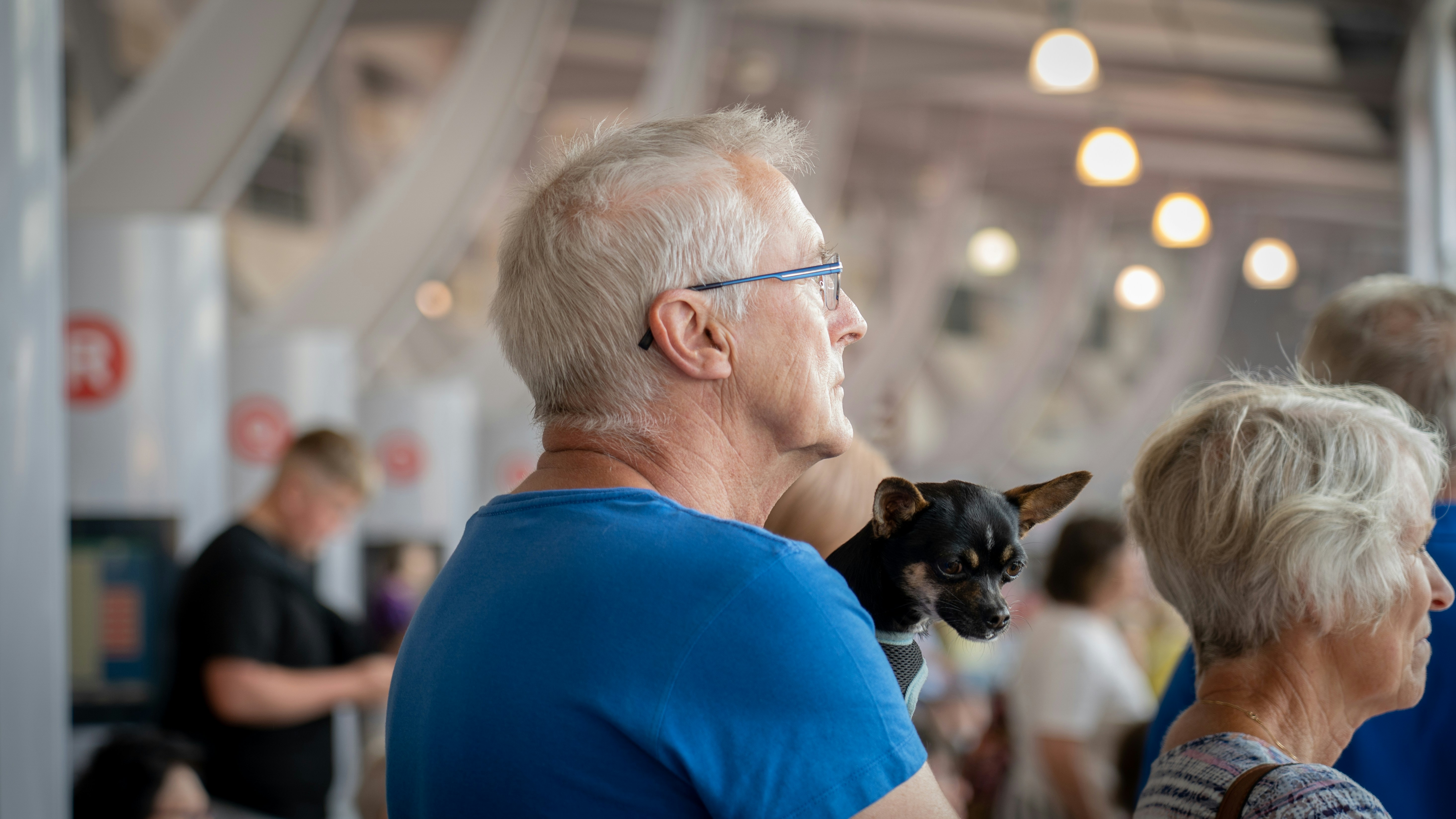 Man in a blue shirt holding a small dog in a busy indoor setting with soft lighting.