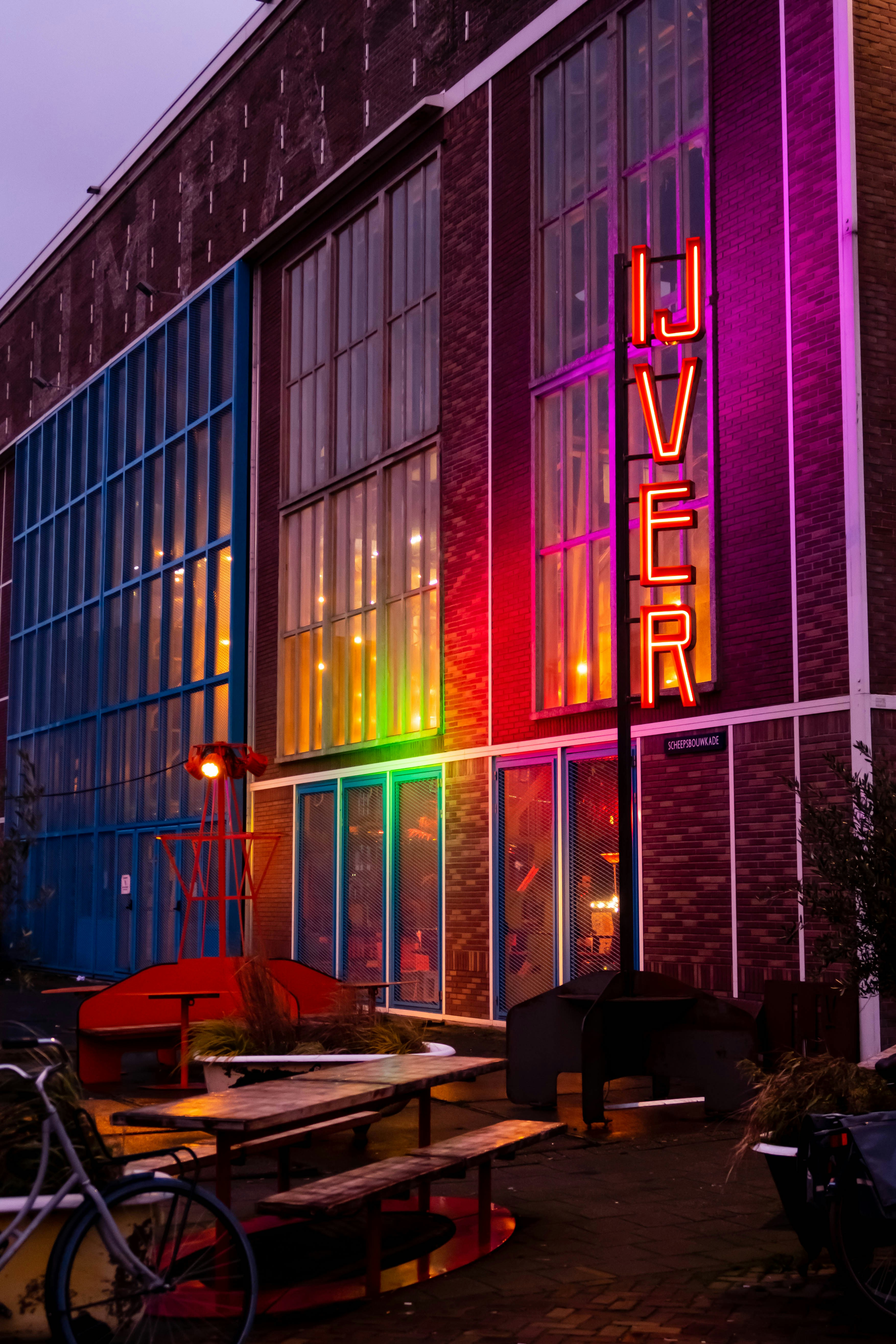 Vibrant neon sign reading 'IJVER' illuminated against a brick building, showcasing a lively urban atmosphere with colorful reflections on the glass windows.