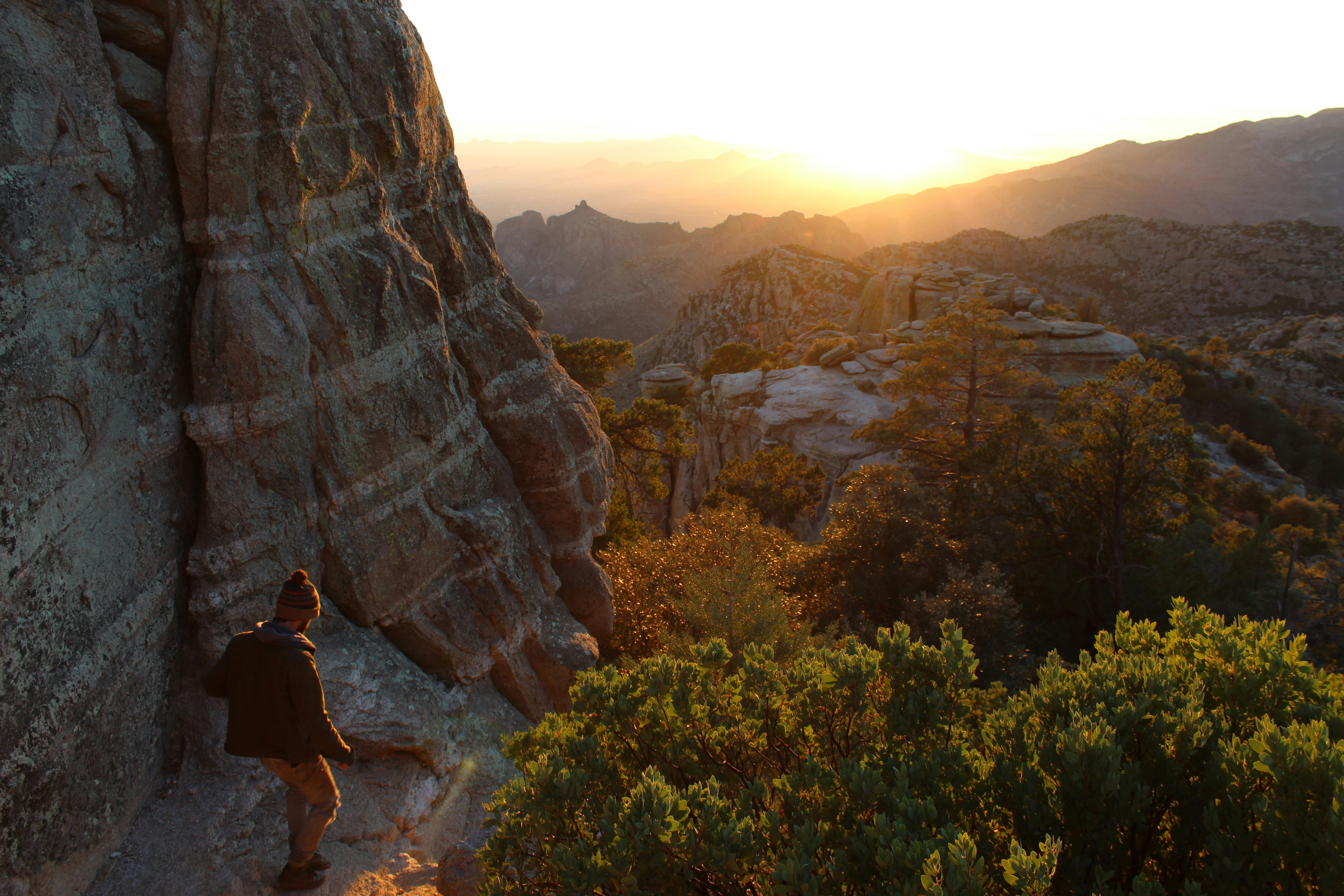 A hiker navigates rocky terrain as the sun sets behind distant mountains, casting a warm glow over the landscape.